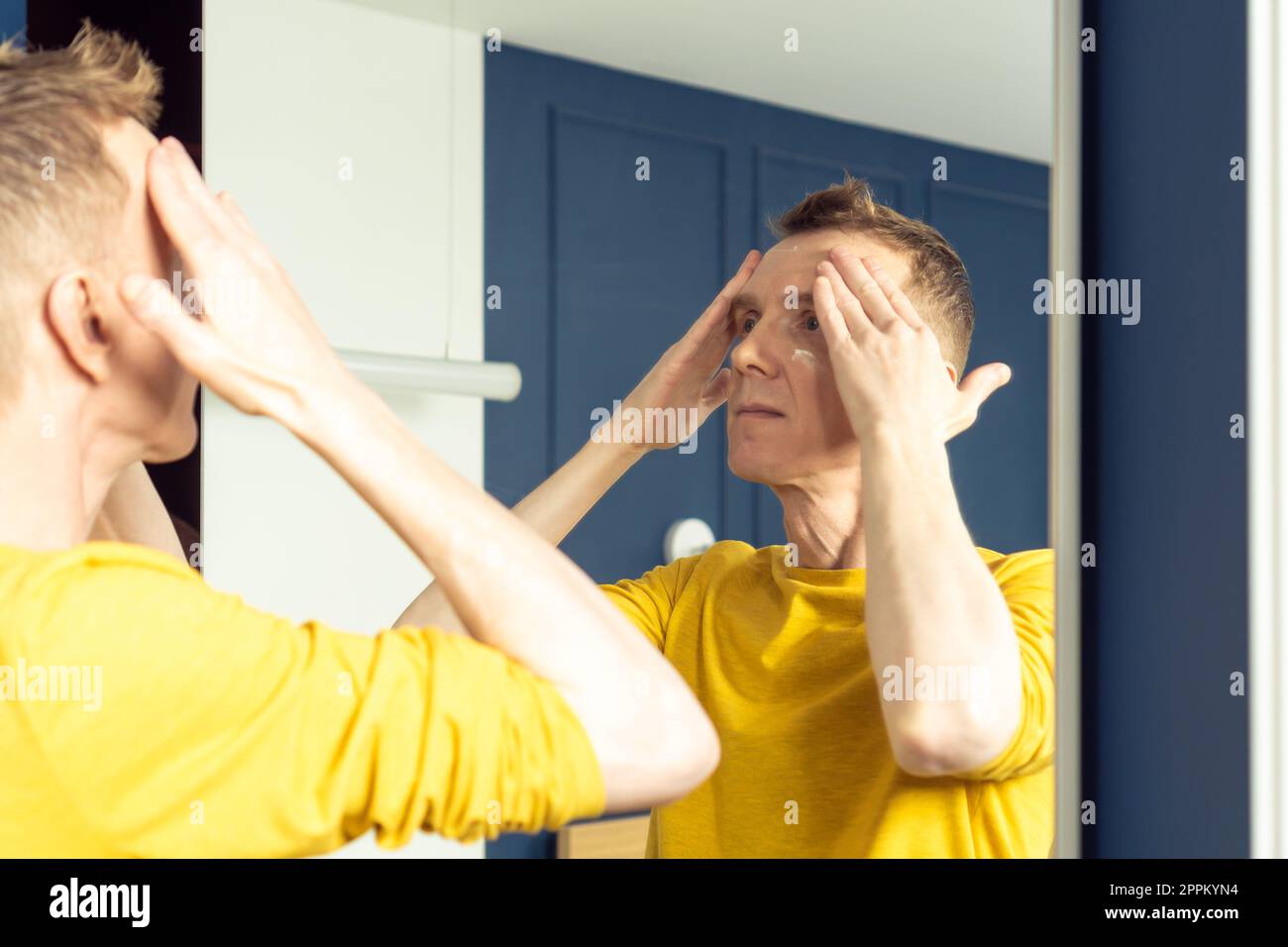 Middle aged man apply cream on clear face, then massage with hands. Male portrait in mirror ...