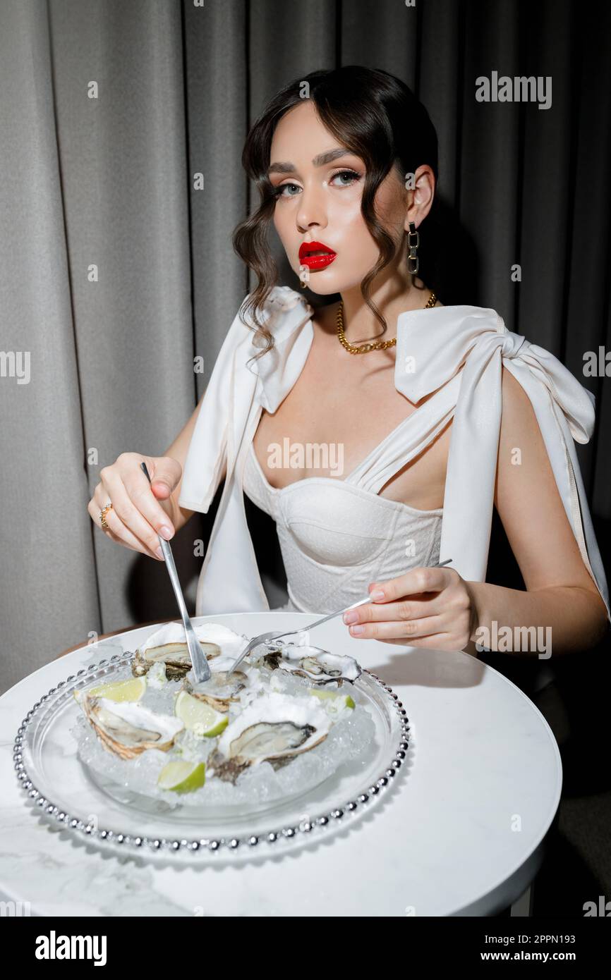 A young European girl with red lips in a cream dress eats oysters in a ...