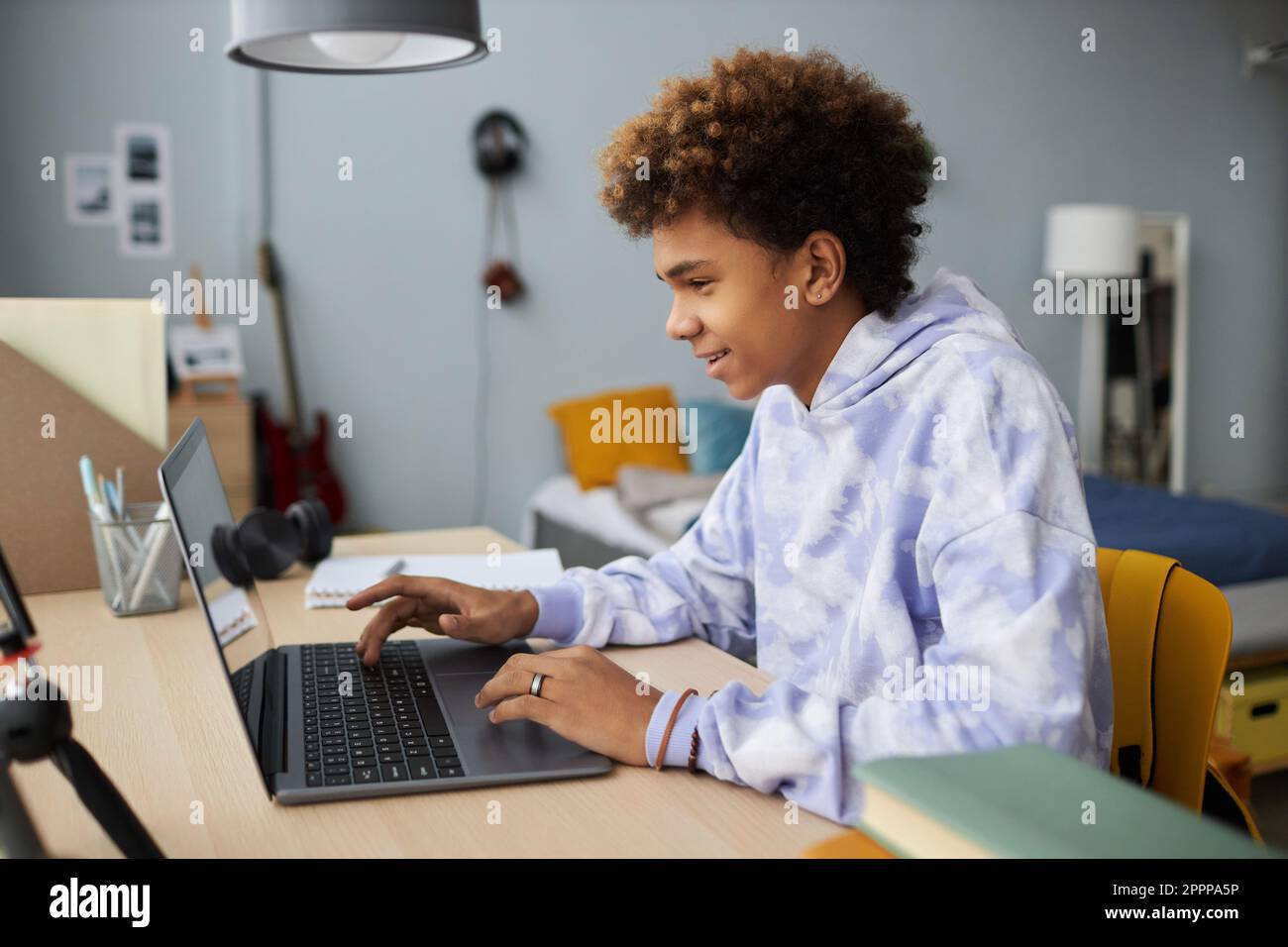 Smiling teenage boy in hoodie typing on laptop keyboard and looking at ...