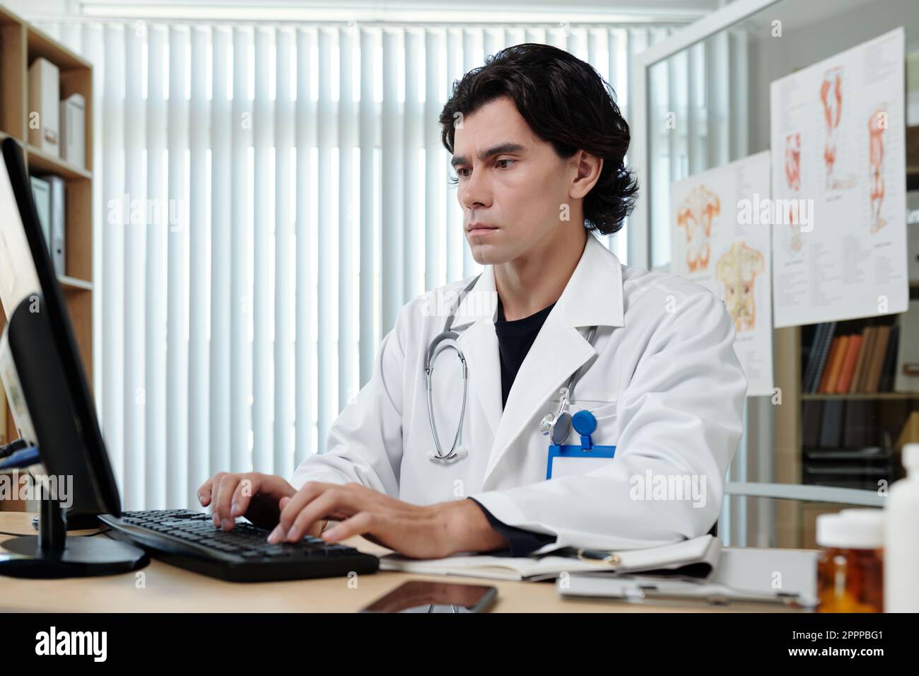 Young serious doctor in lab coat typing on keyboard while sitting by ...