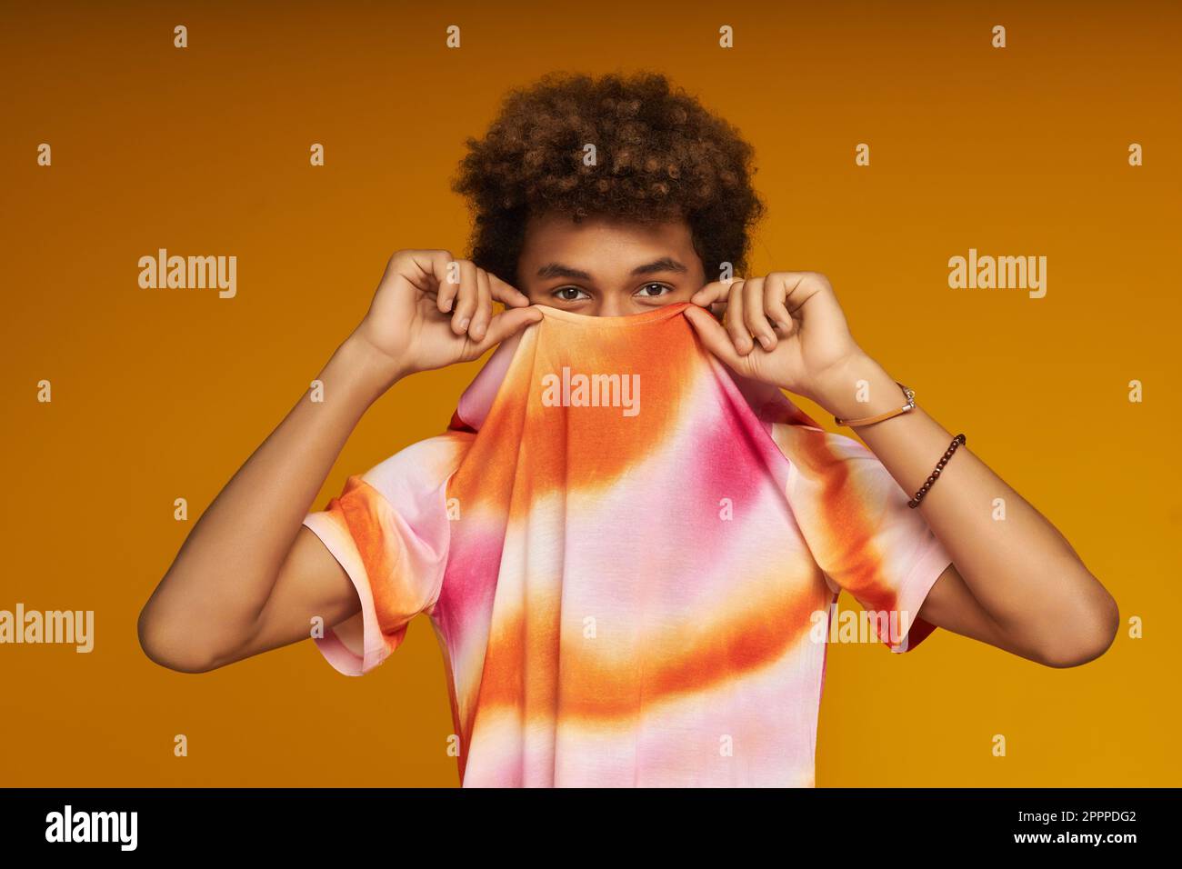 Cute teenage boy hiding face behind colorful t-shirt while standing in ...