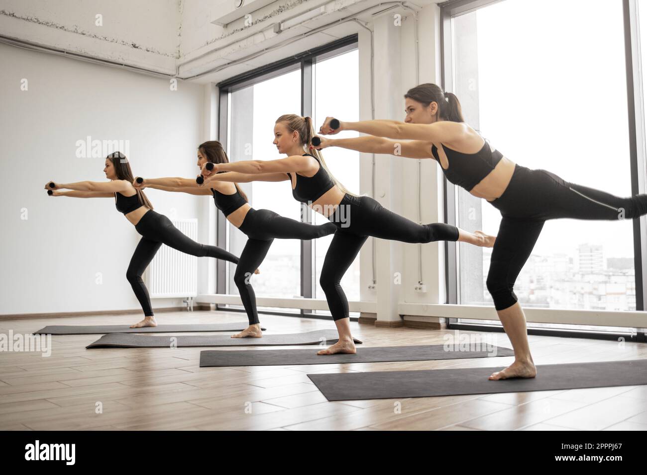 Group of active women in sportswear balancing in Warrior III Pose with dumbbells in hands while ...