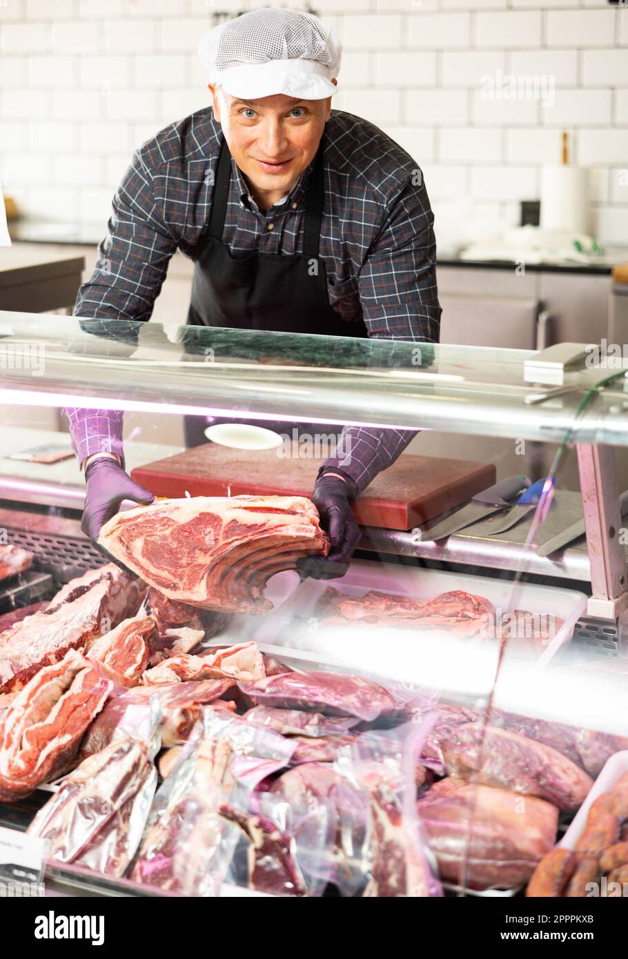 Positive male butcher holding big chunk of beef ribs in meat section of ...