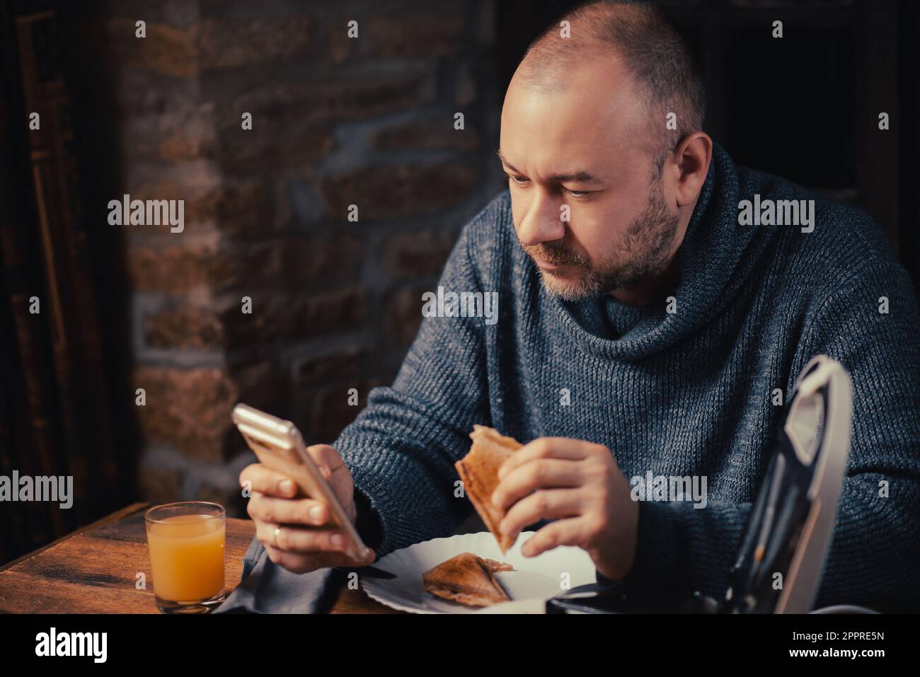 A man eats a sandwich and looks at the phone at home. Ordinary middle ...