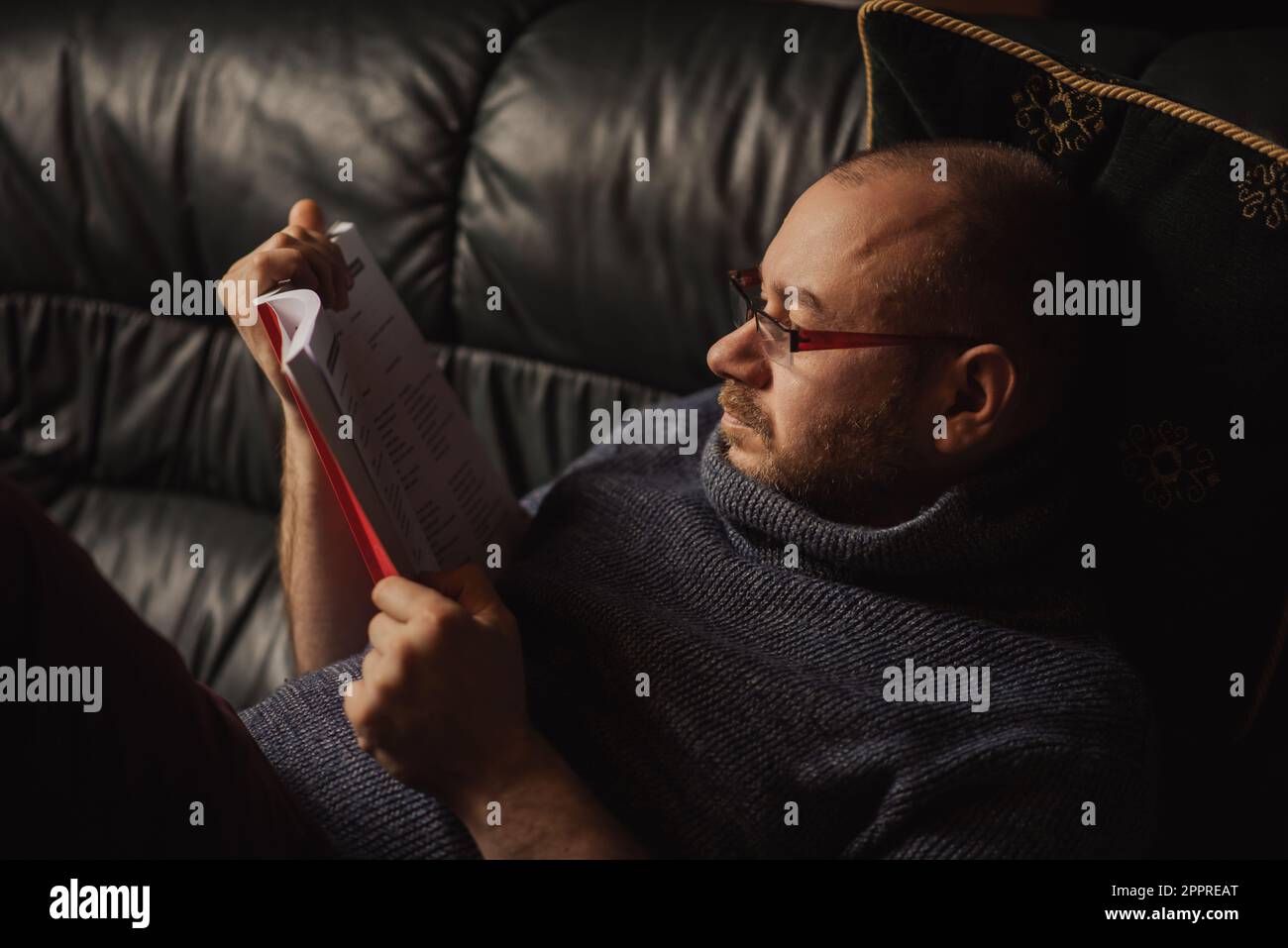 A man with glasses is reading a book while lying on the couch. Middle ...
