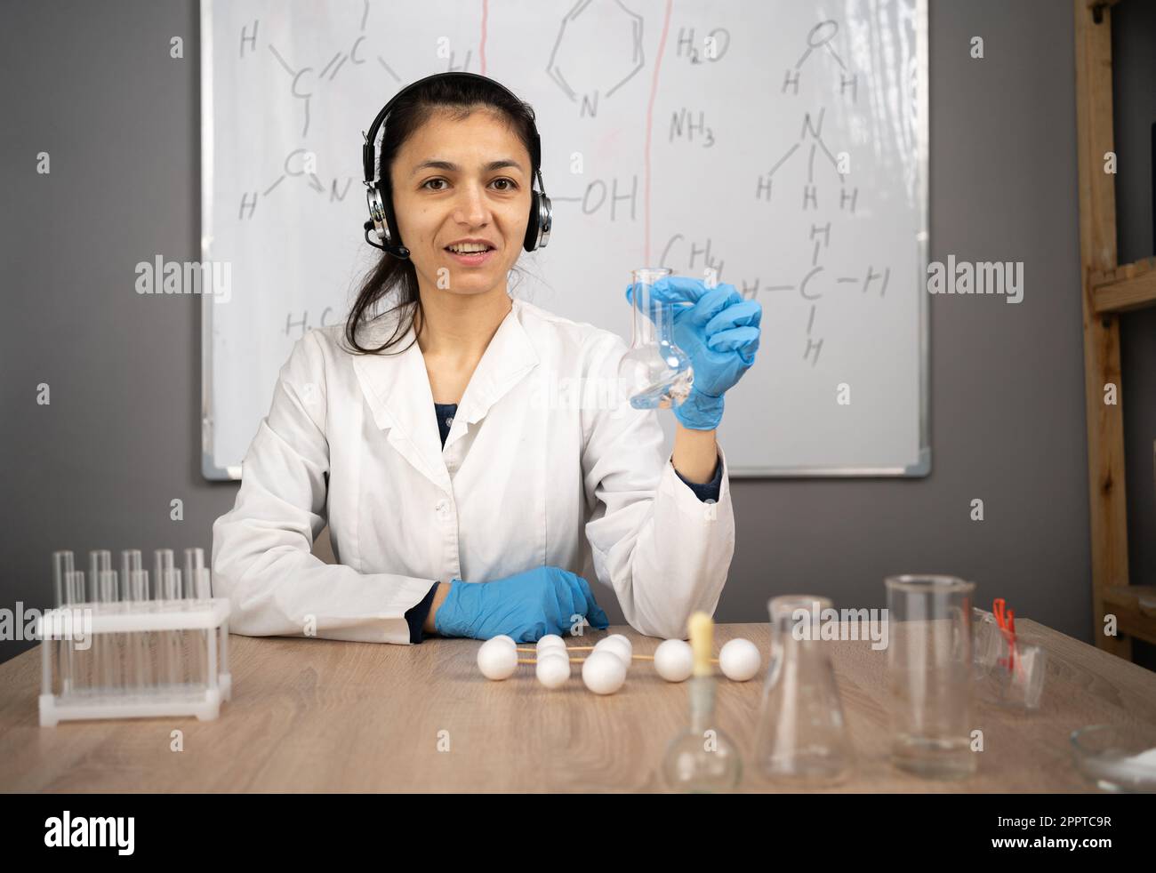Smiling female chemistry teacher with headset having online class and showing experiments in ...