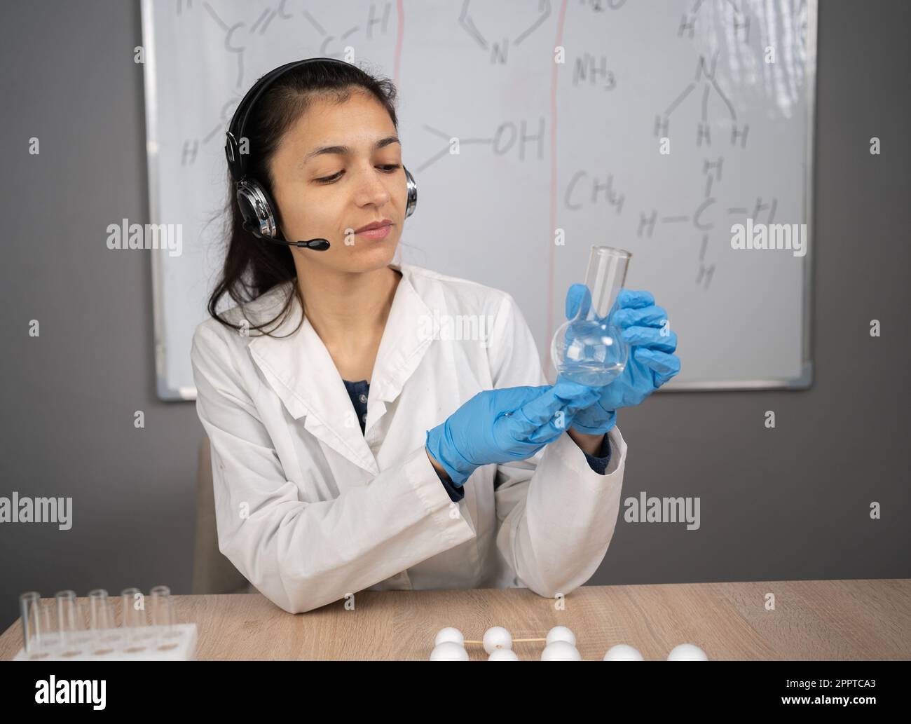 Smiling female chemistry teacher with headset having online class and showing experiments in ...