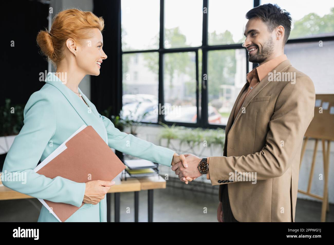side view of pleased businesswoman with folder shaking hands with young ...