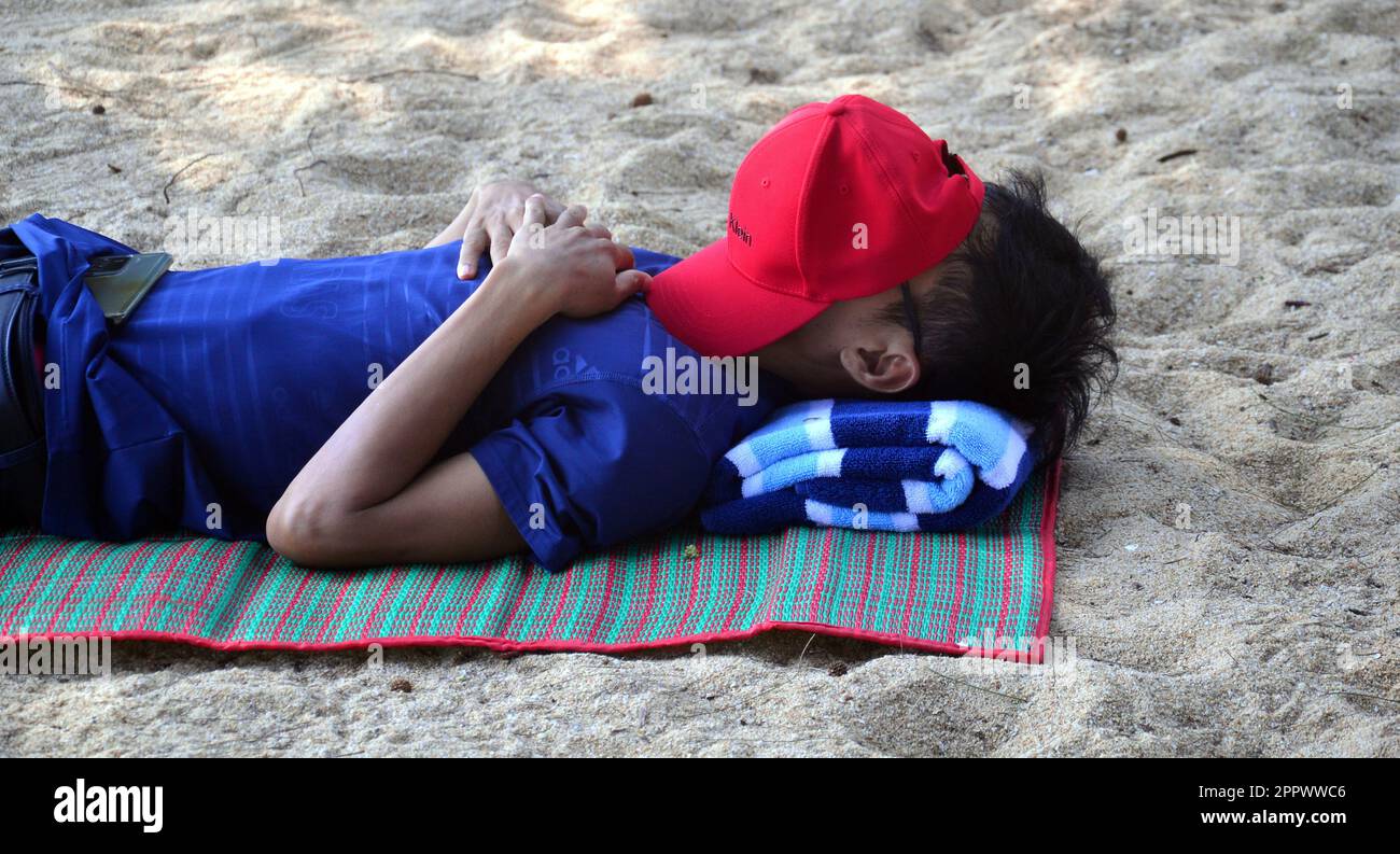 A man with a baseball cap covering his face lies on the beach in ...