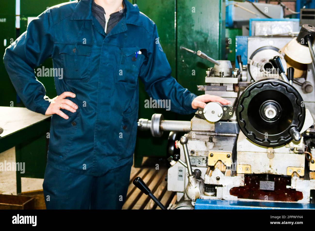 A man working in a robe, overalls stands next to an industrial lathe ...
