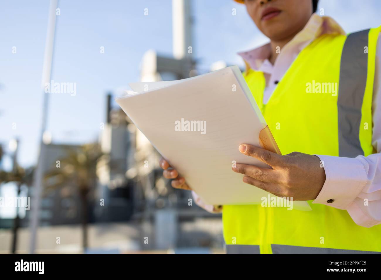 Young cropped unrecognizable woman in hardhat and engineer vest reading ...