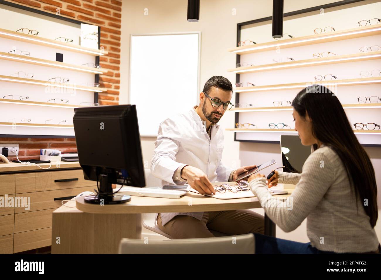 Male optician in white uniform assisting woman in selecting eyeglasses ...