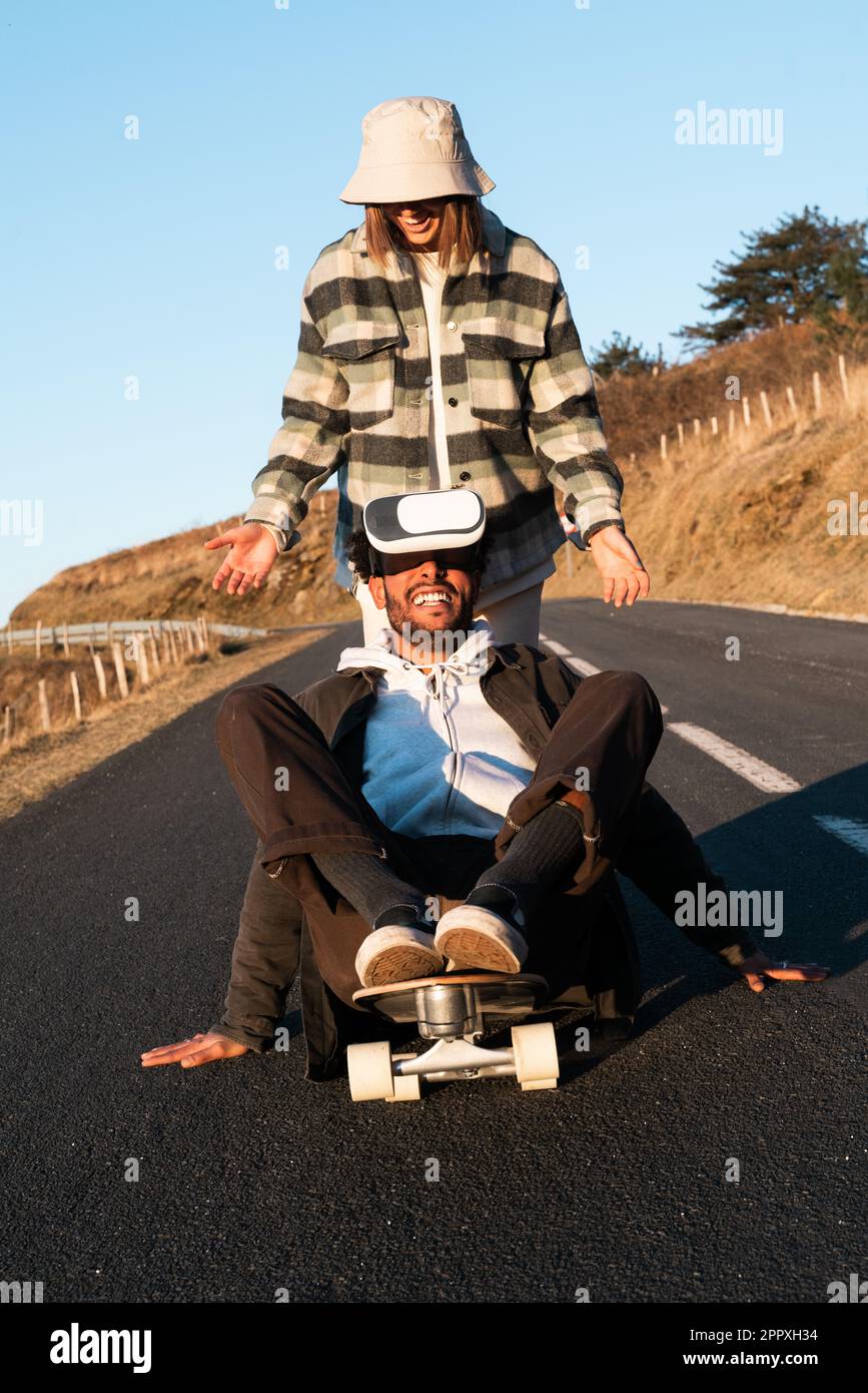 Full body of young male with VR goggles sitting on skateboard while ...