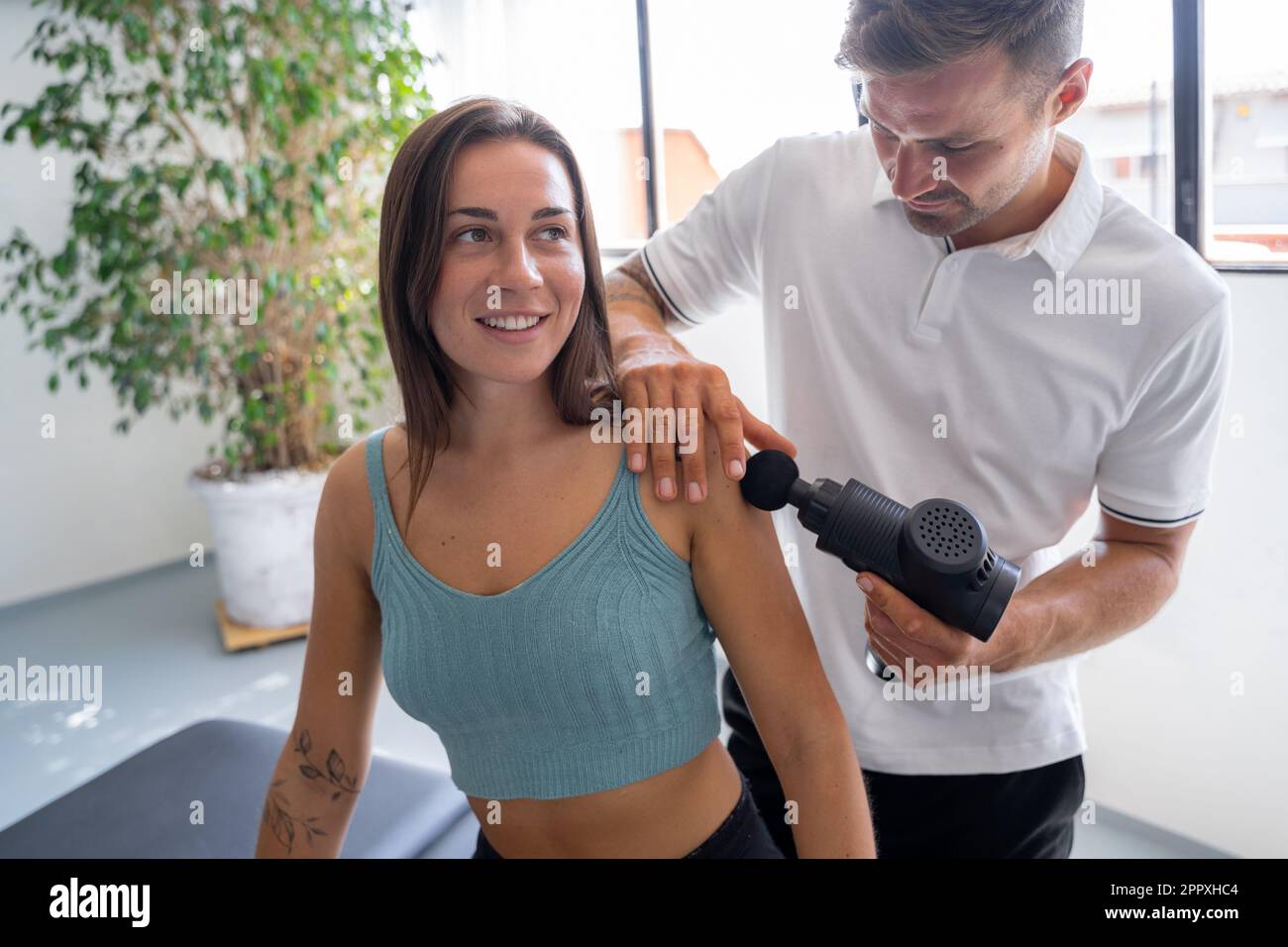 Young Concentrated Male Therapist Using Percussion Gun On Shoulder Of Female Client Sitting On