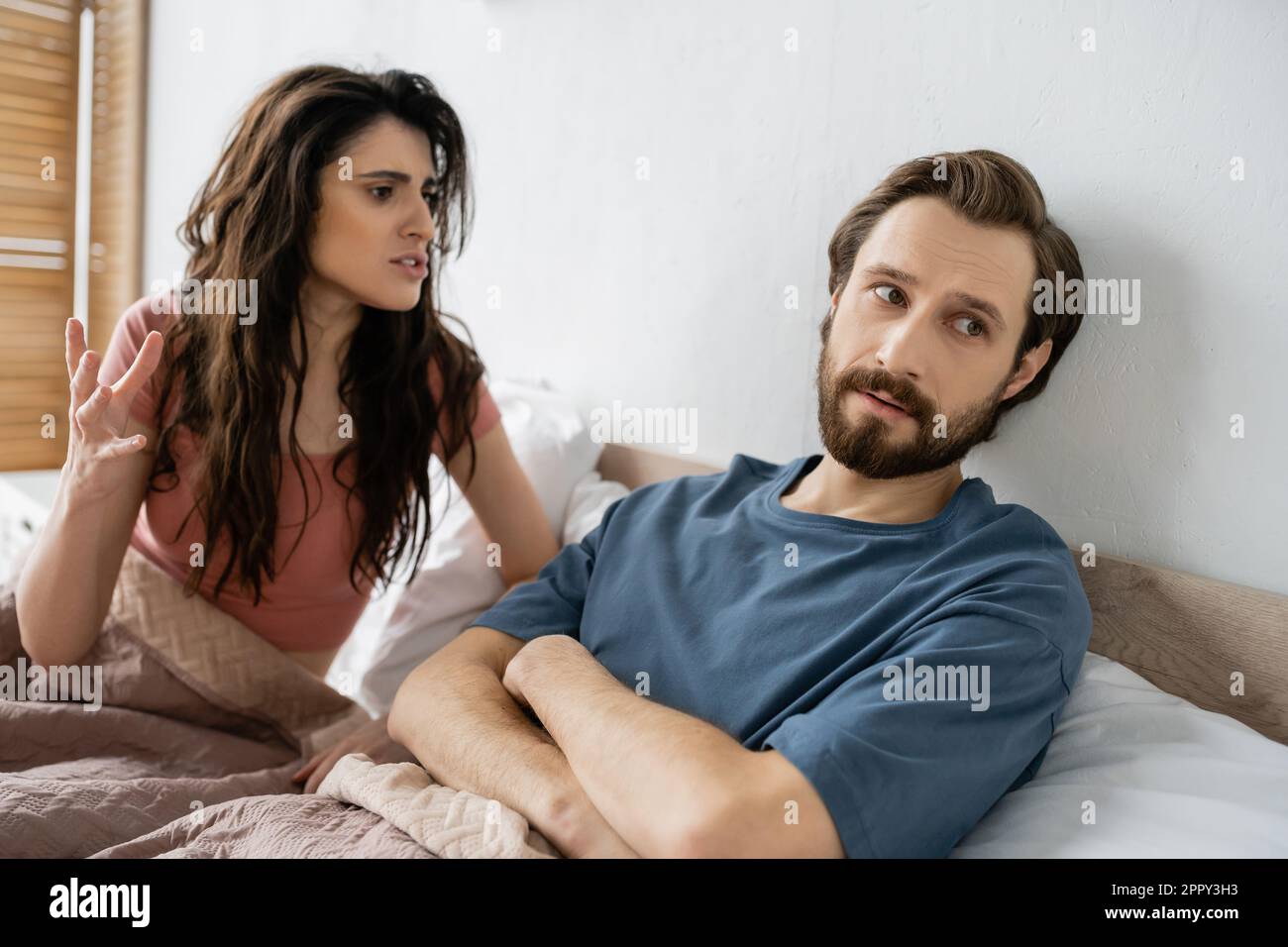 Angry woman talking to bearded boyfriend crossing arms on bed at home ...