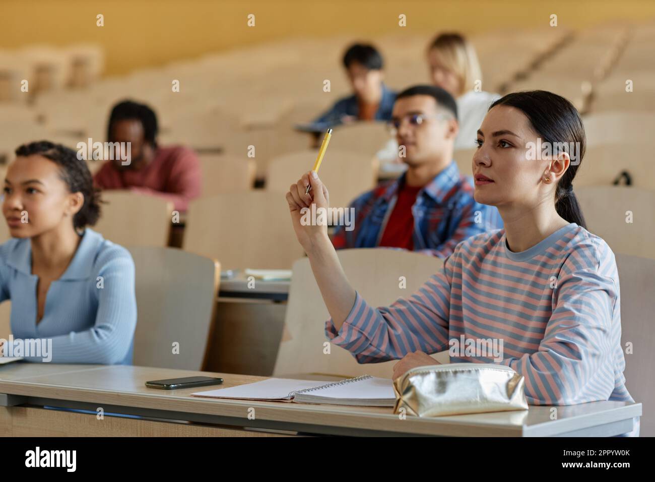 Student raising her arm to ask question while sitting at desk with her ...