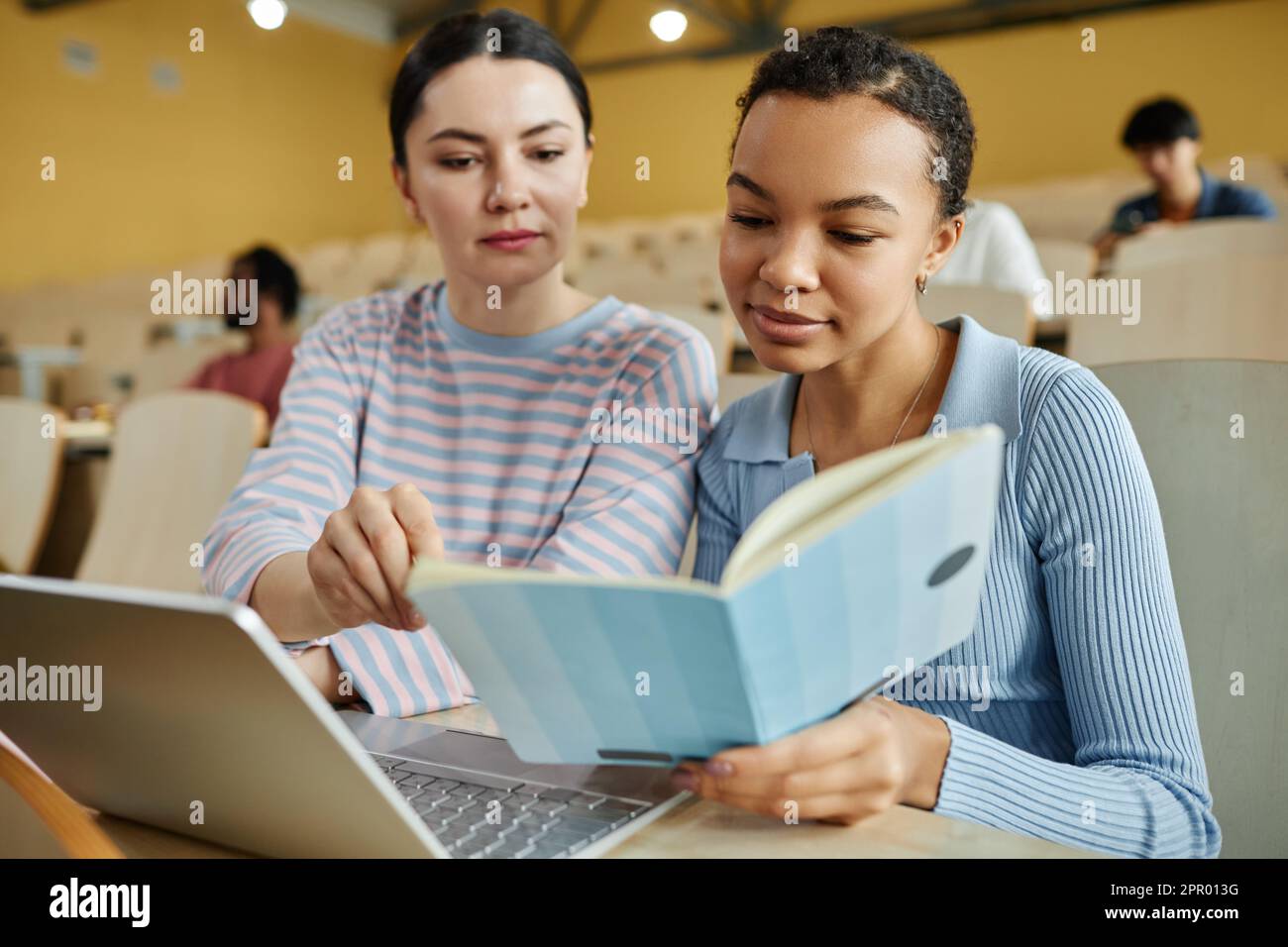 Two girls reading textbook and using laptop during teamwork at lecture ...