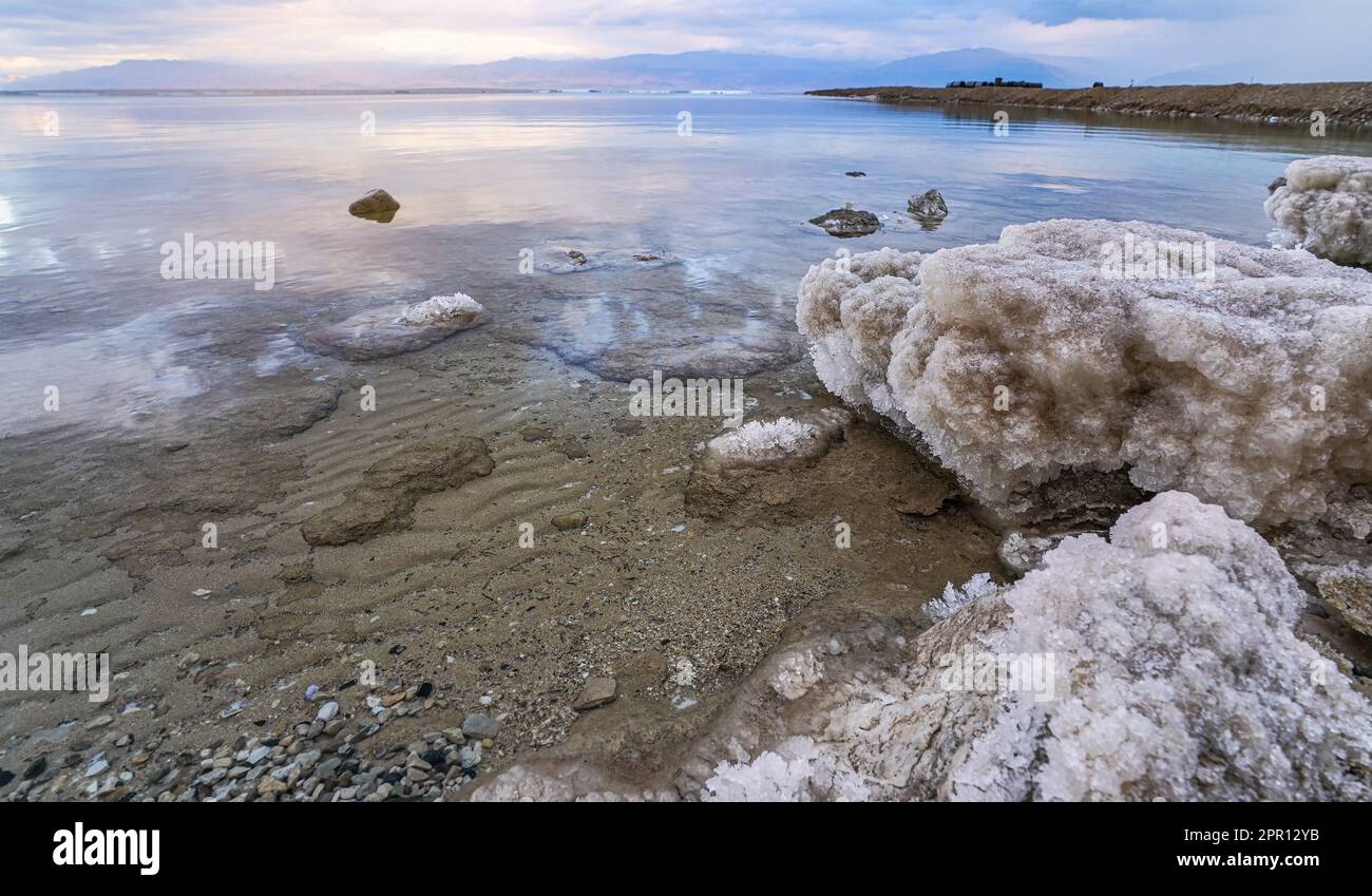 Sand and stones covered with crystalline salt on shore of Dead Sea, clear water near - typical ...