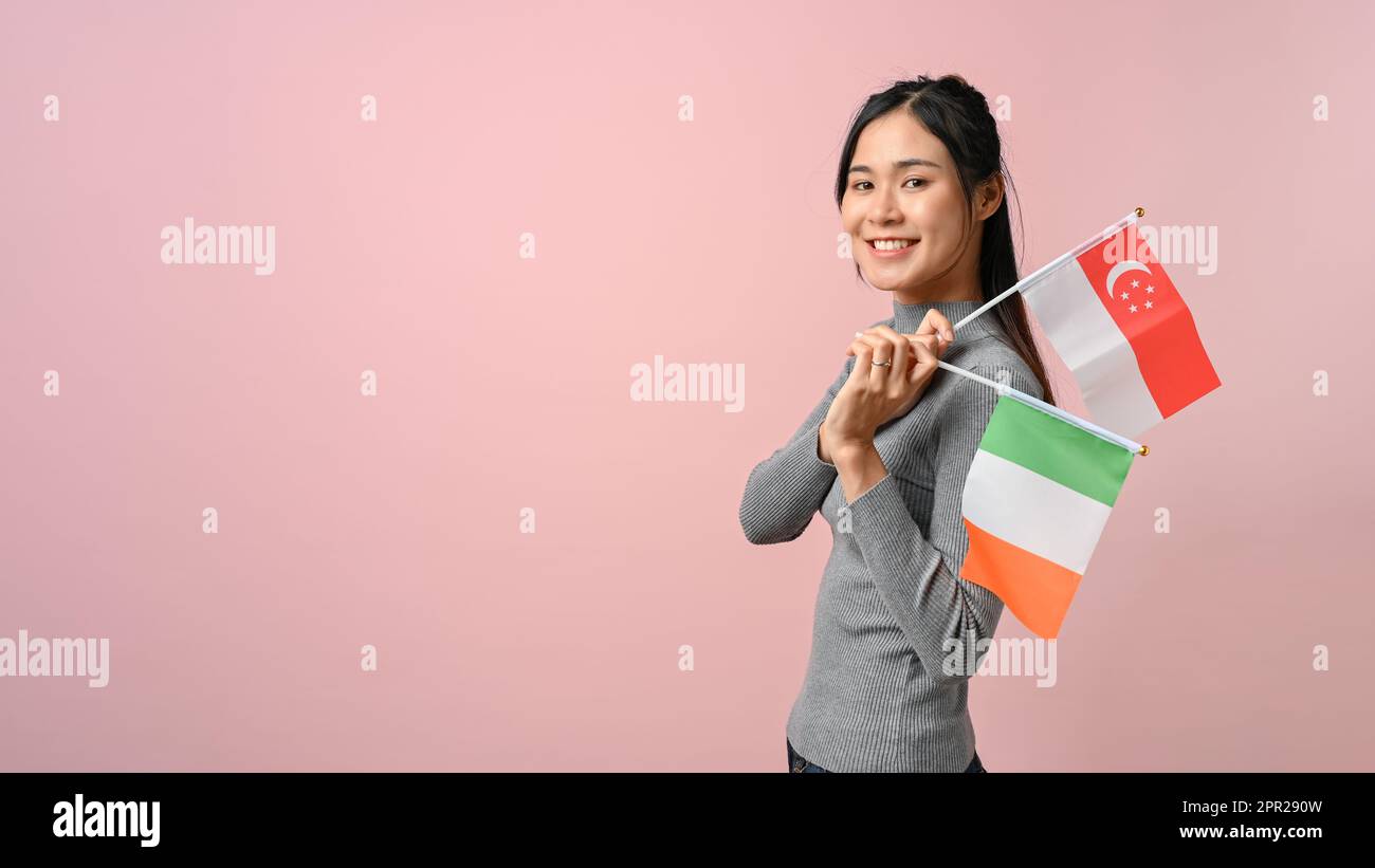 Cheerful young Asian woman showing a Singapore and Ireland flag on pink ...