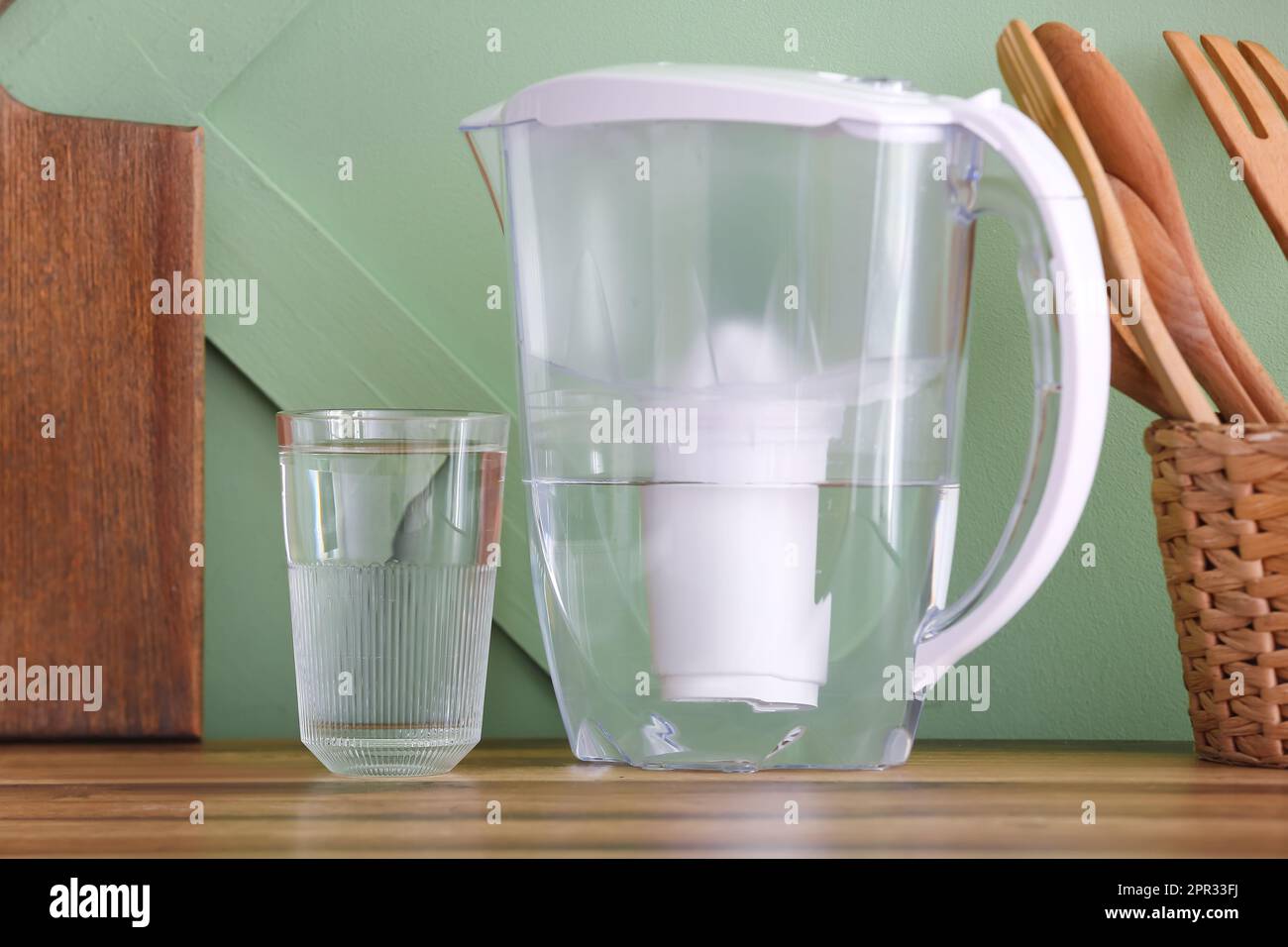 Modern filter jug and glass of water on kitchen counter near green wall ...