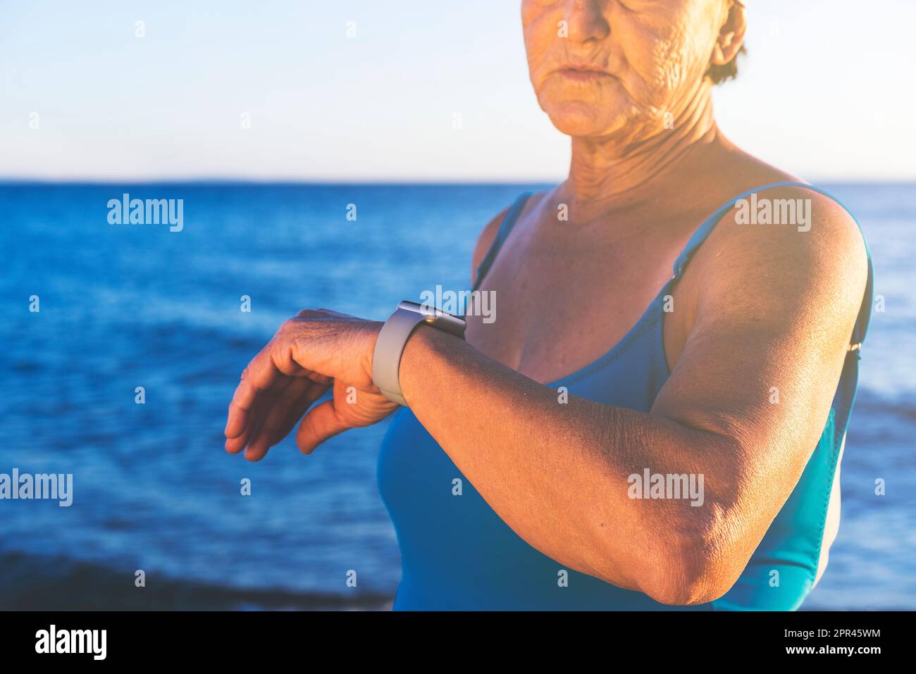 Senior woman at the beach, standing by the sea. Wearing a blue swimsuit ...