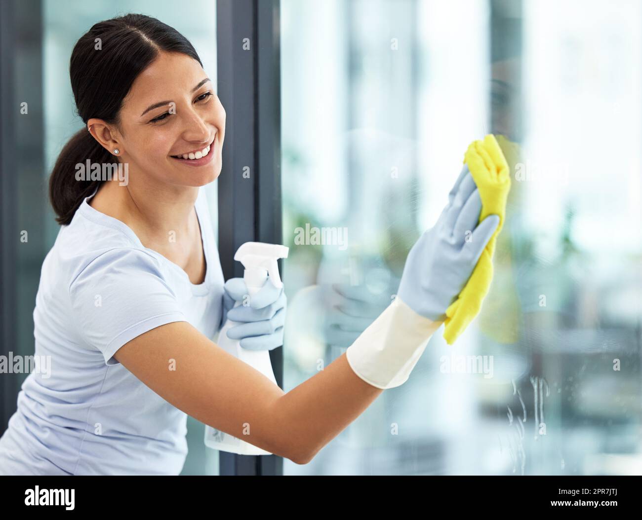 A happy smiling mixed race domestic worker using a cloth on a window ...