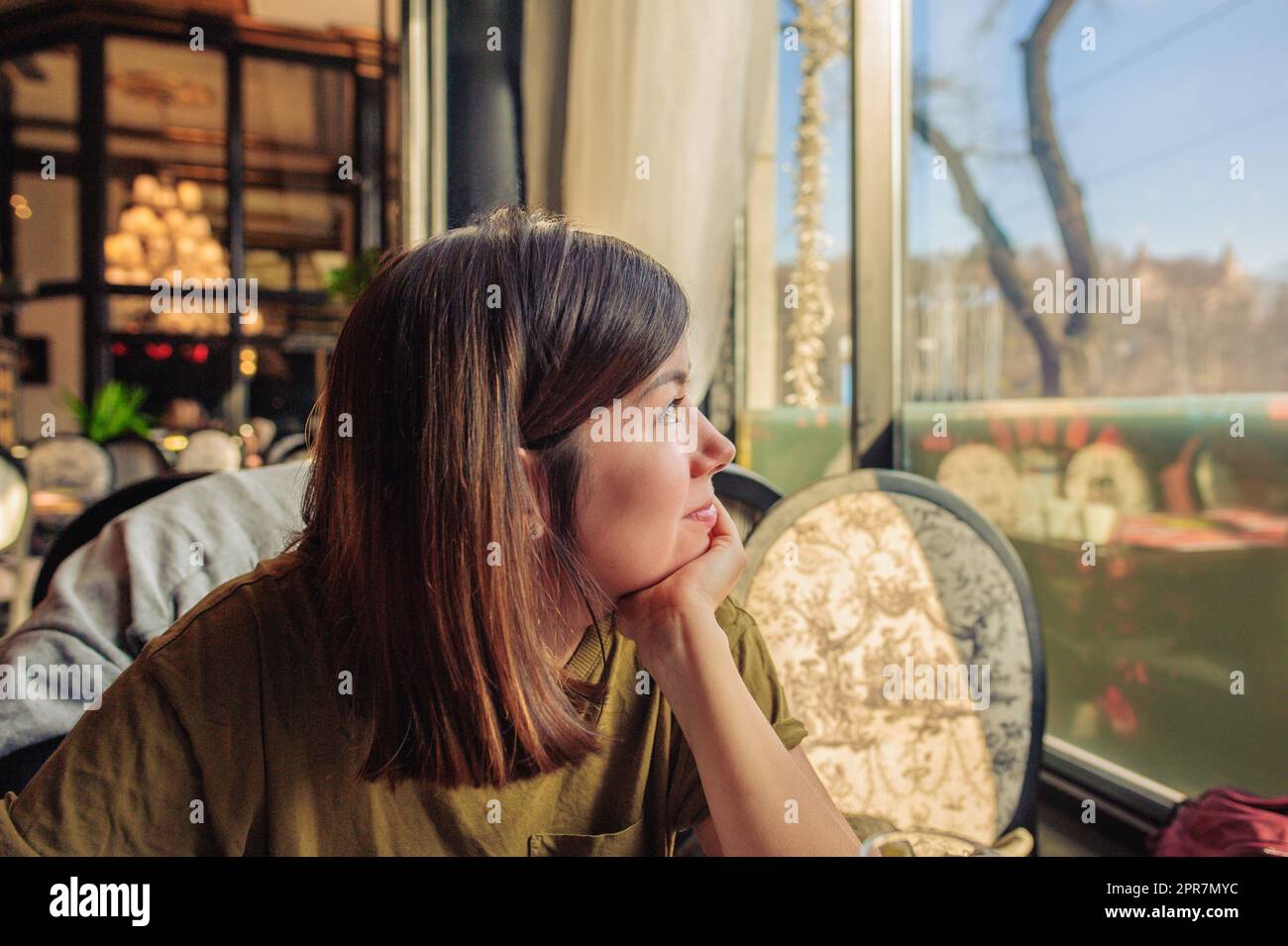 A beautiful young girl sits in a restaurant and looks out the window ...