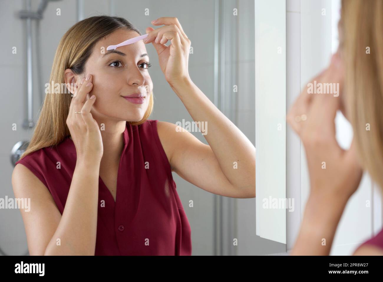 Hispanic woman shaving her eyebrows with razor in the mirror at home ...