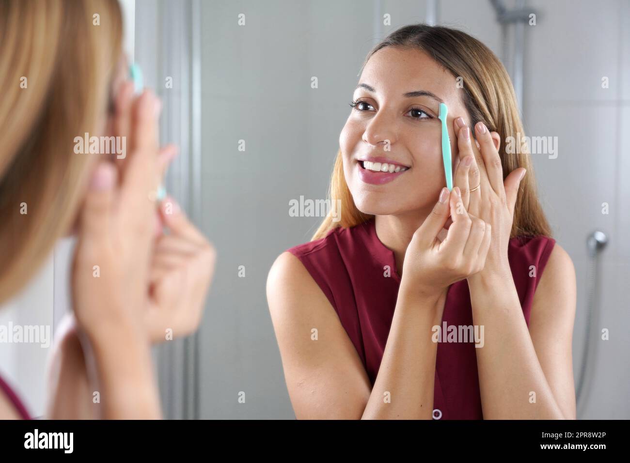 Beautiful young woman shaving her eyebrows by razor on the mirror at ...