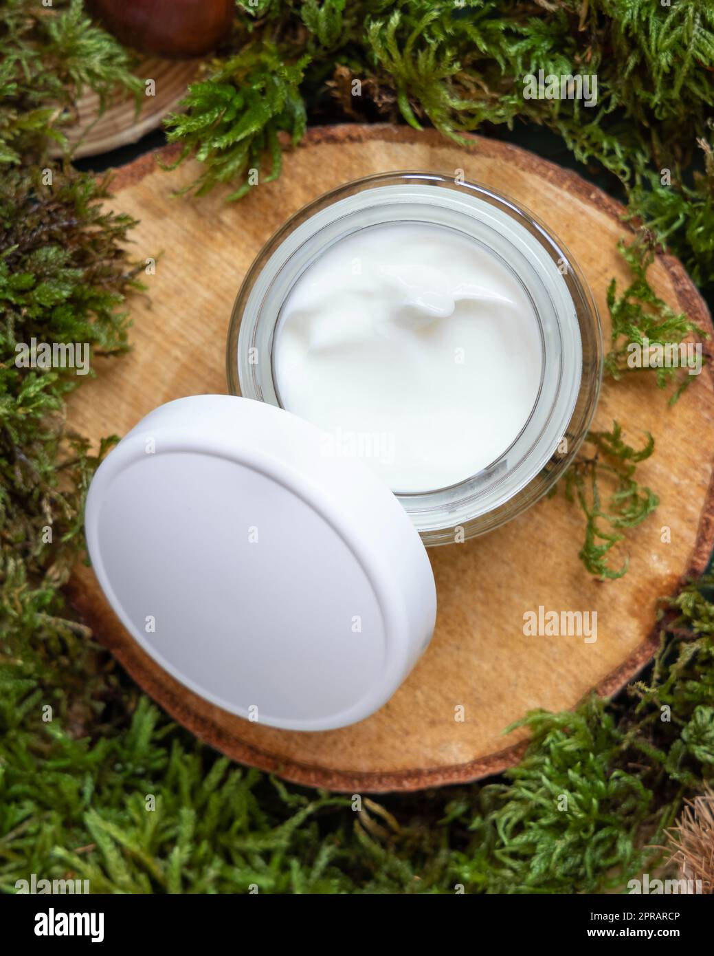 Opened cream jar with white cap on wooden piece between green moss, top ...