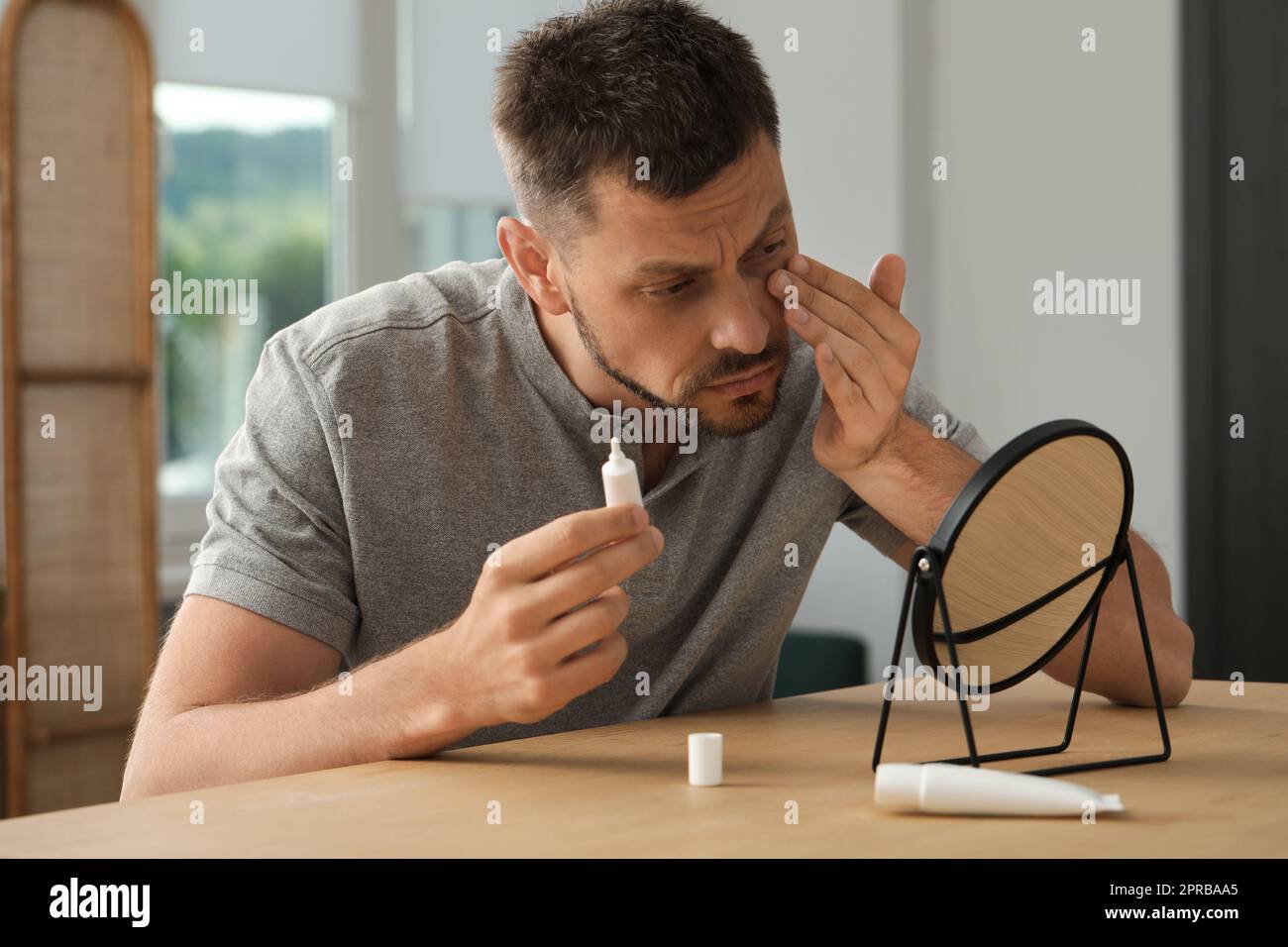 Sleep deprived man covering up dark circles with concealer near mirror ...