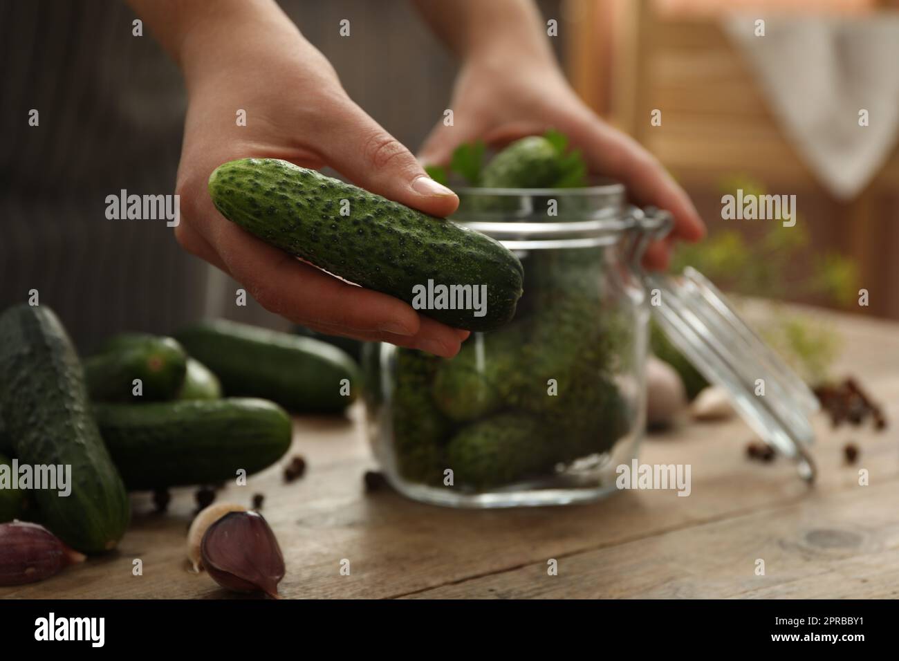 Woman putting cucumber into glass jar at wooden kitchen table, closeup ...