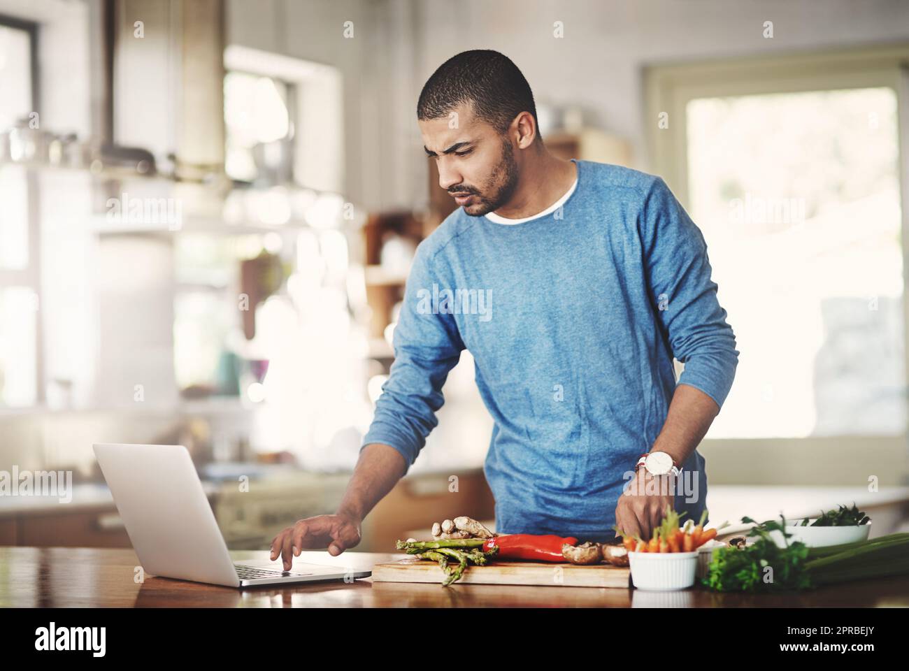 Young man using a laptop while cooking in a kitchen, checking the ...