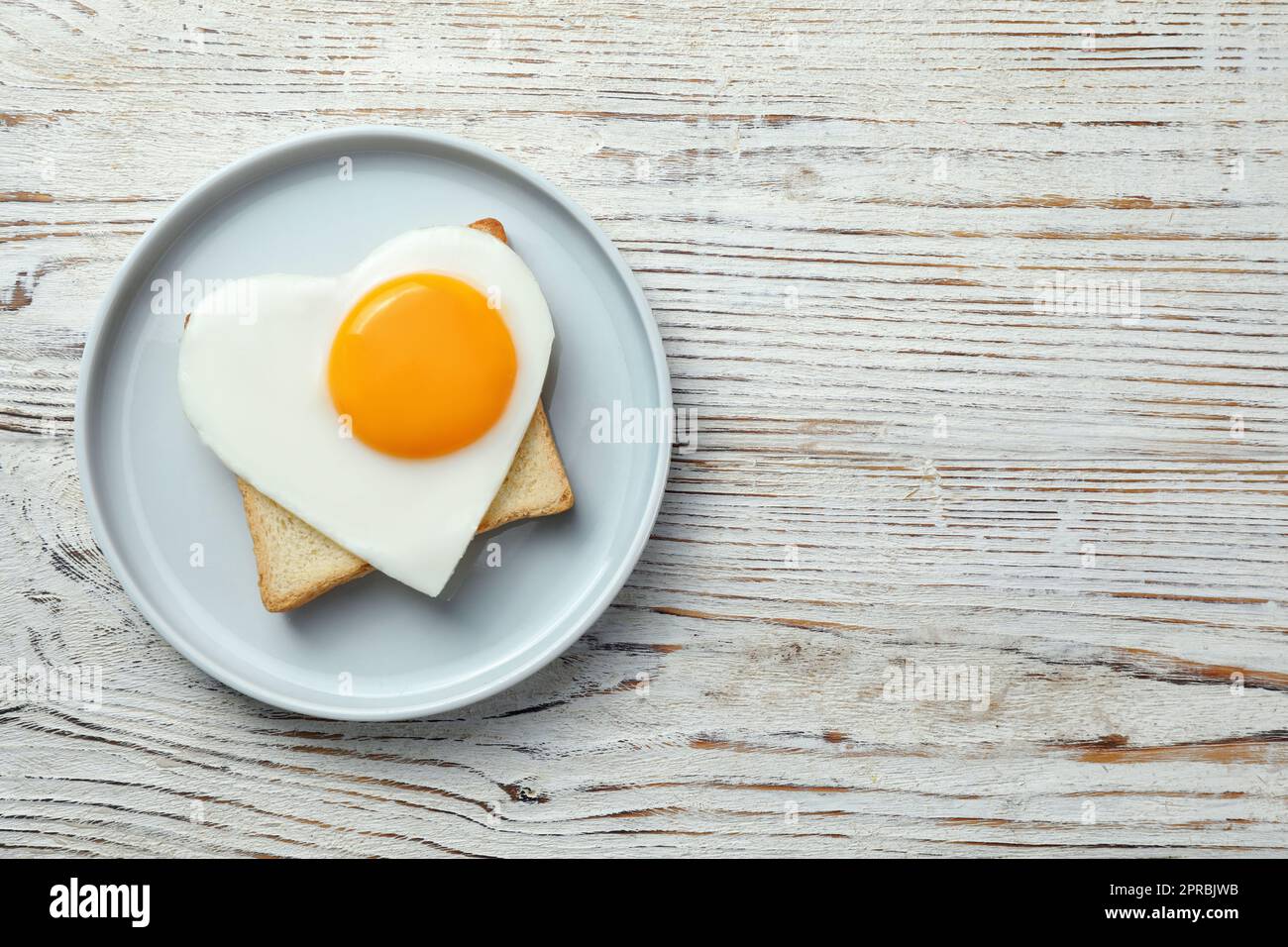 Romantic breakfast with heart shaped fried egg and toast on white wooden table, top view. Space ...