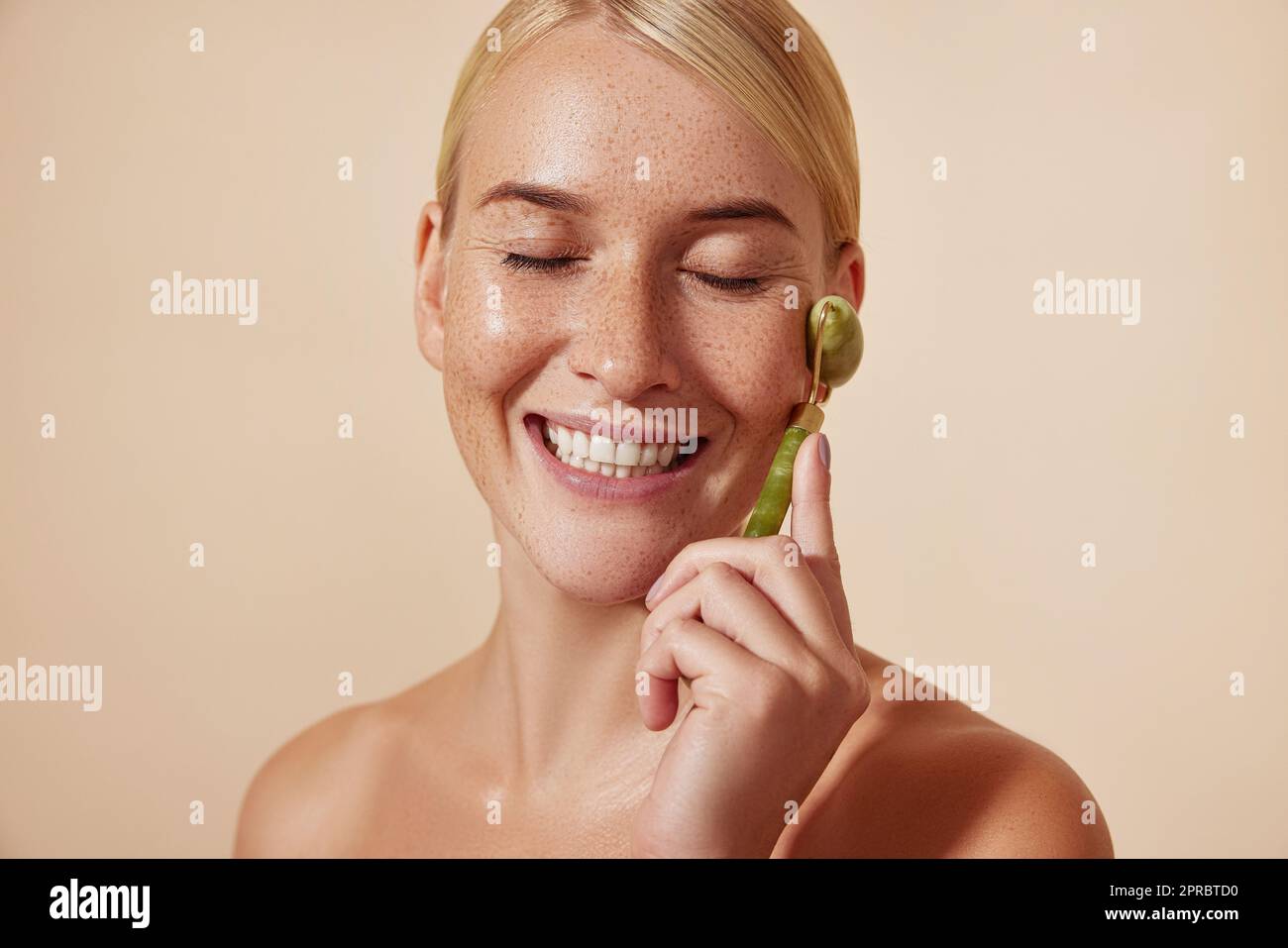 Happy woman with freckles and smooth skin using a jade stone roller ...