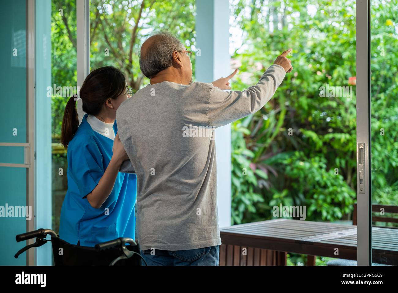 Young Asian nurse is supporting elderly man sitting in wheelchair walking to looking out window ...