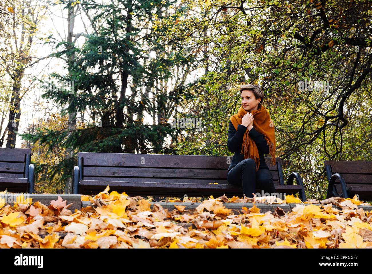 A short-haired woman wraps herself in a scarf sitting on a bench in an ...