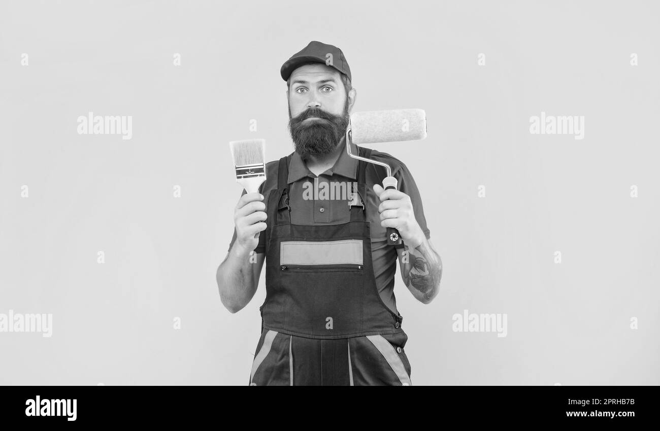 bearded puzzled man painter in work clothes hold paint roller and brush ...