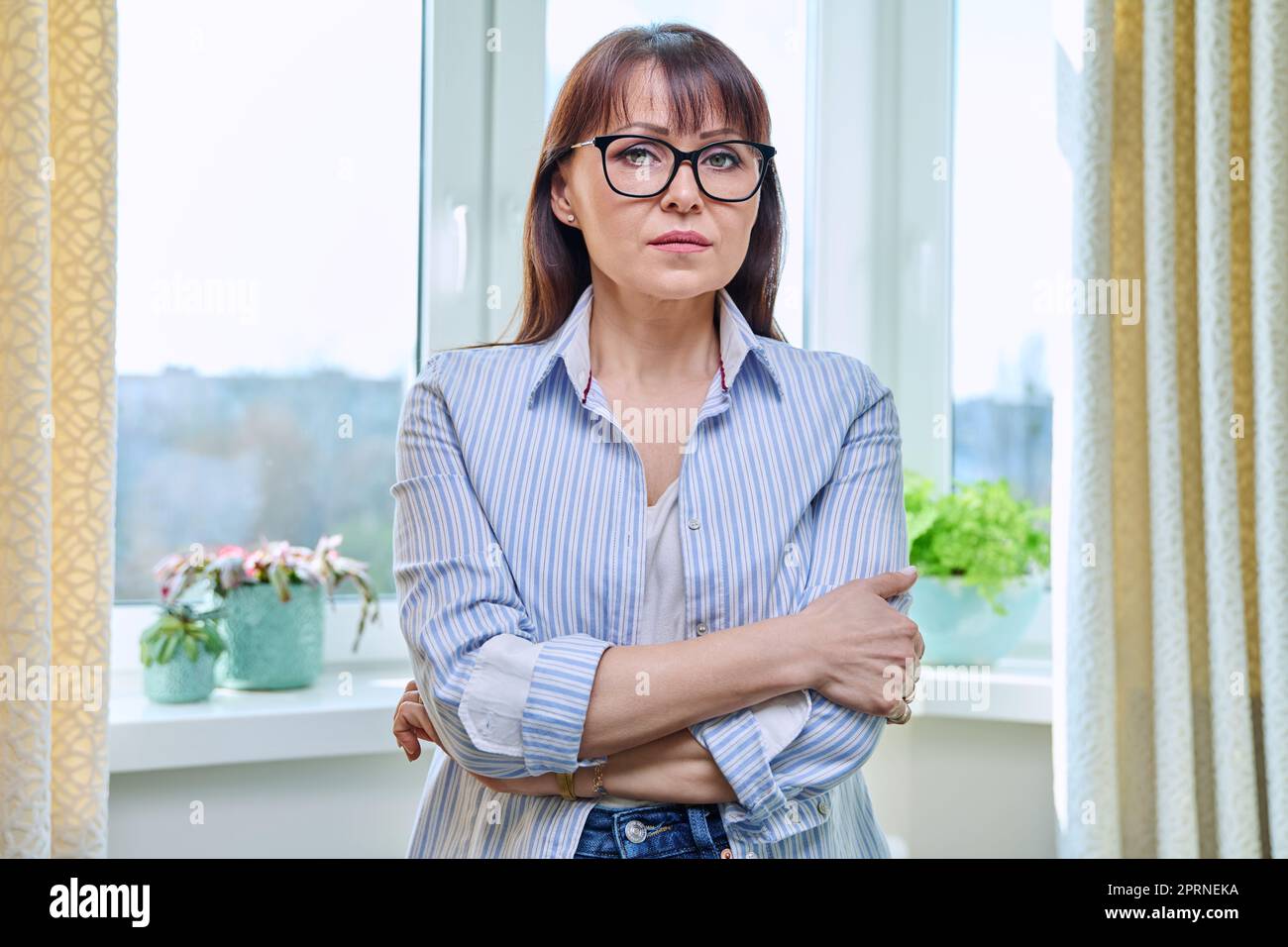 Portrait of confident serious mature woman posing near window at home ...