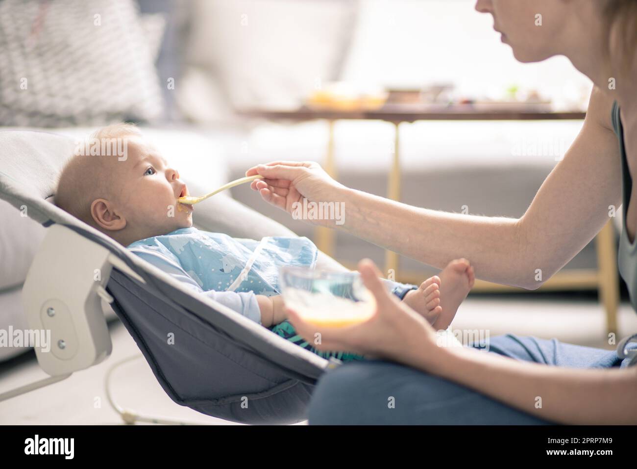 Mother spoon feeding her baby boy child in baby chair with fruit puree ...