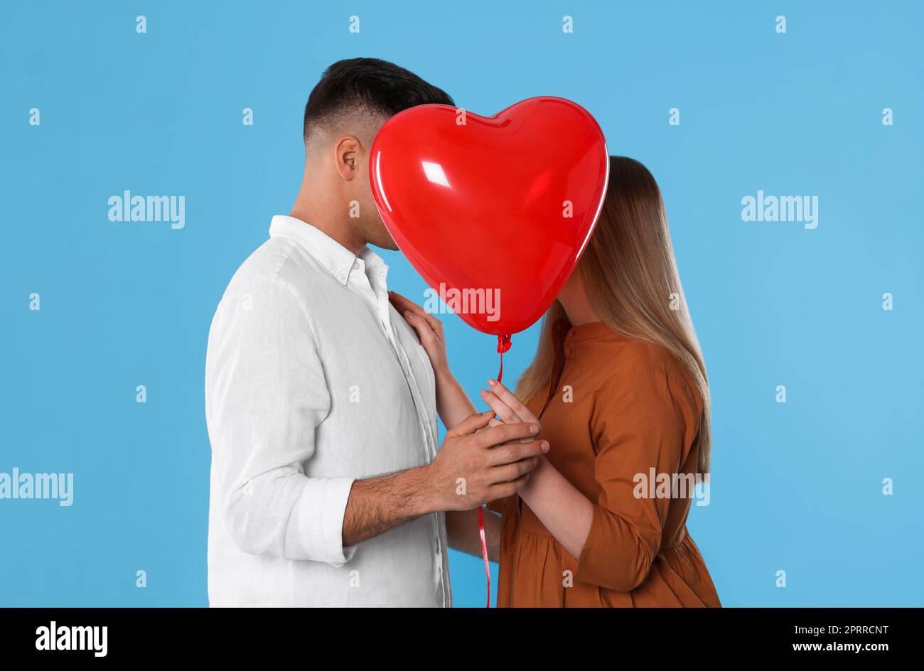 Lovely couple kissing behind heart shaped balloon on light blue ...