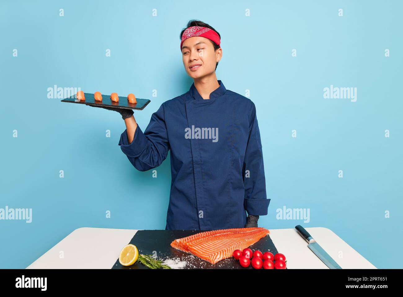 Charming asian man in blue cooking uniform stands at table with fish ...