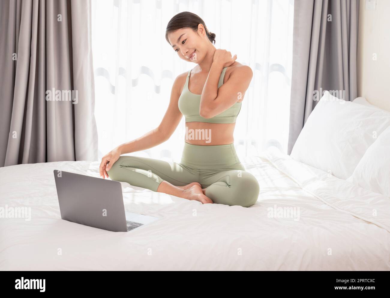Woman practicing yoga on bed after waking up. Asian female sitting on bed using her laptop while ...