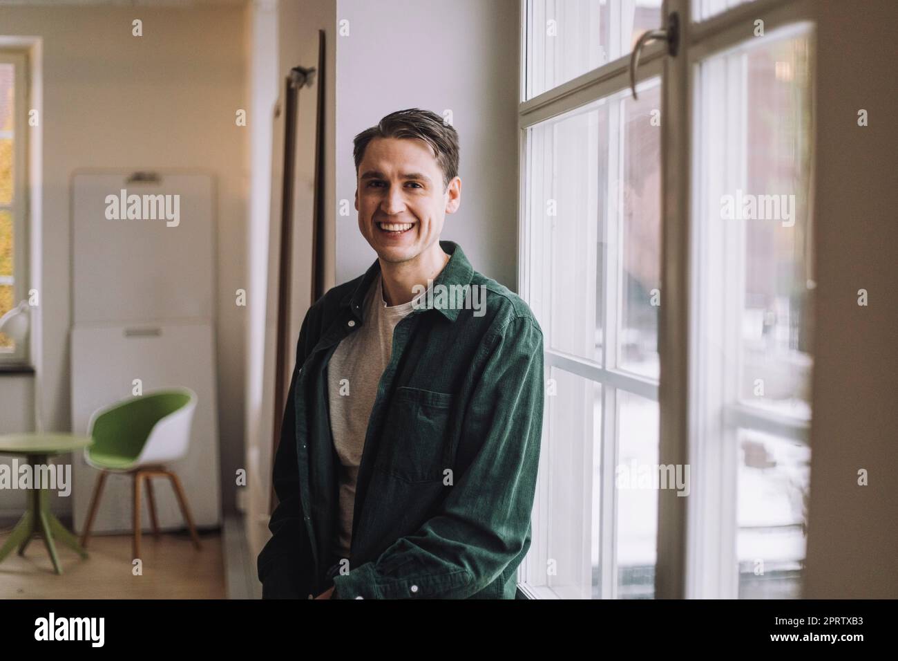 Portrait of happy male PhD student leaning by window in innovation lab ...