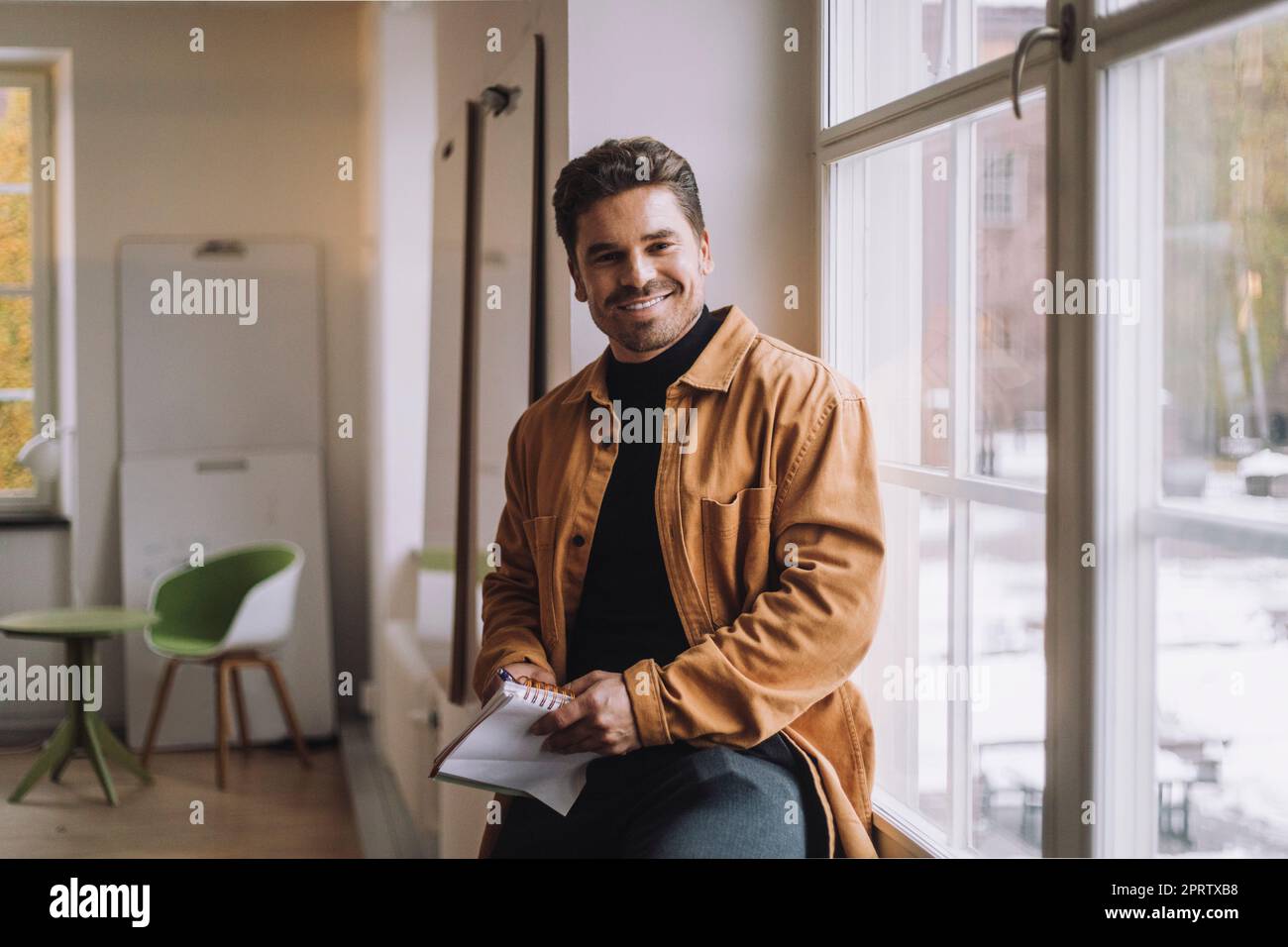 Portrait of smiling male PhD student holding diary sitting by window in ...