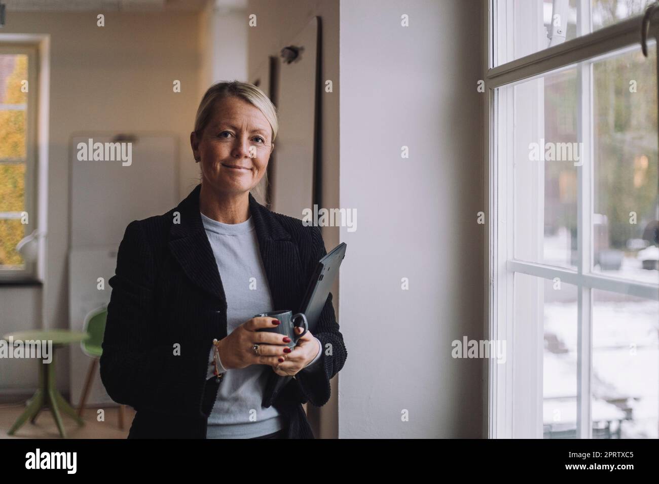 Smiling female professor holding laptop standing by window in ...