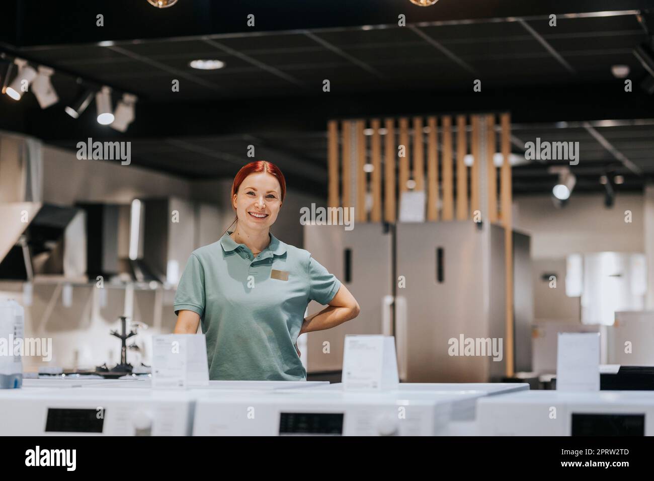 Portrait of smiling female retail clerk standing with hand on hip in ...