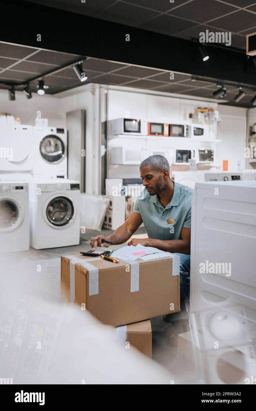 Male sales clerk using calculator on box while working in electronics ...