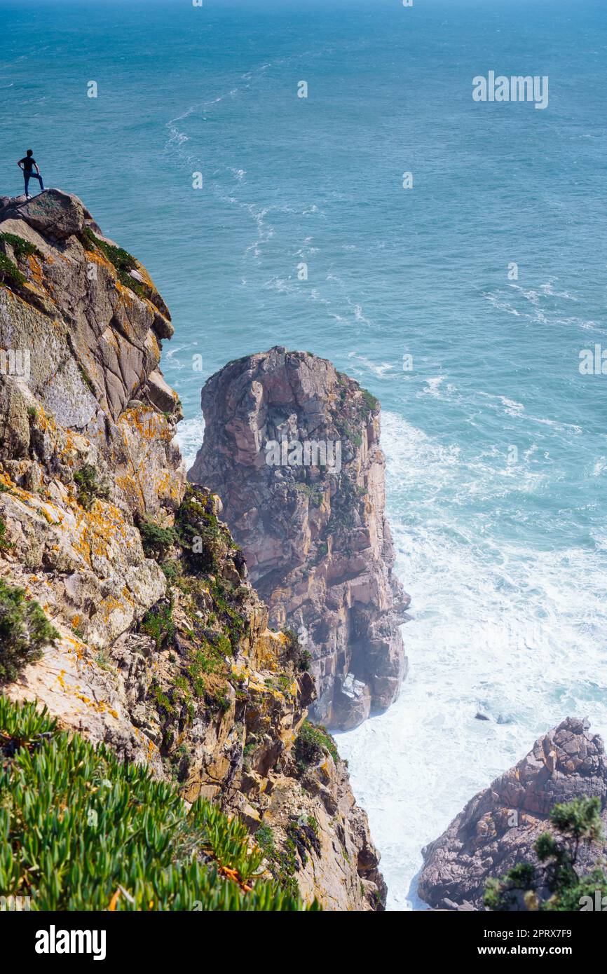 Small human figure stands on top of high cliff, waves are breaking on ...