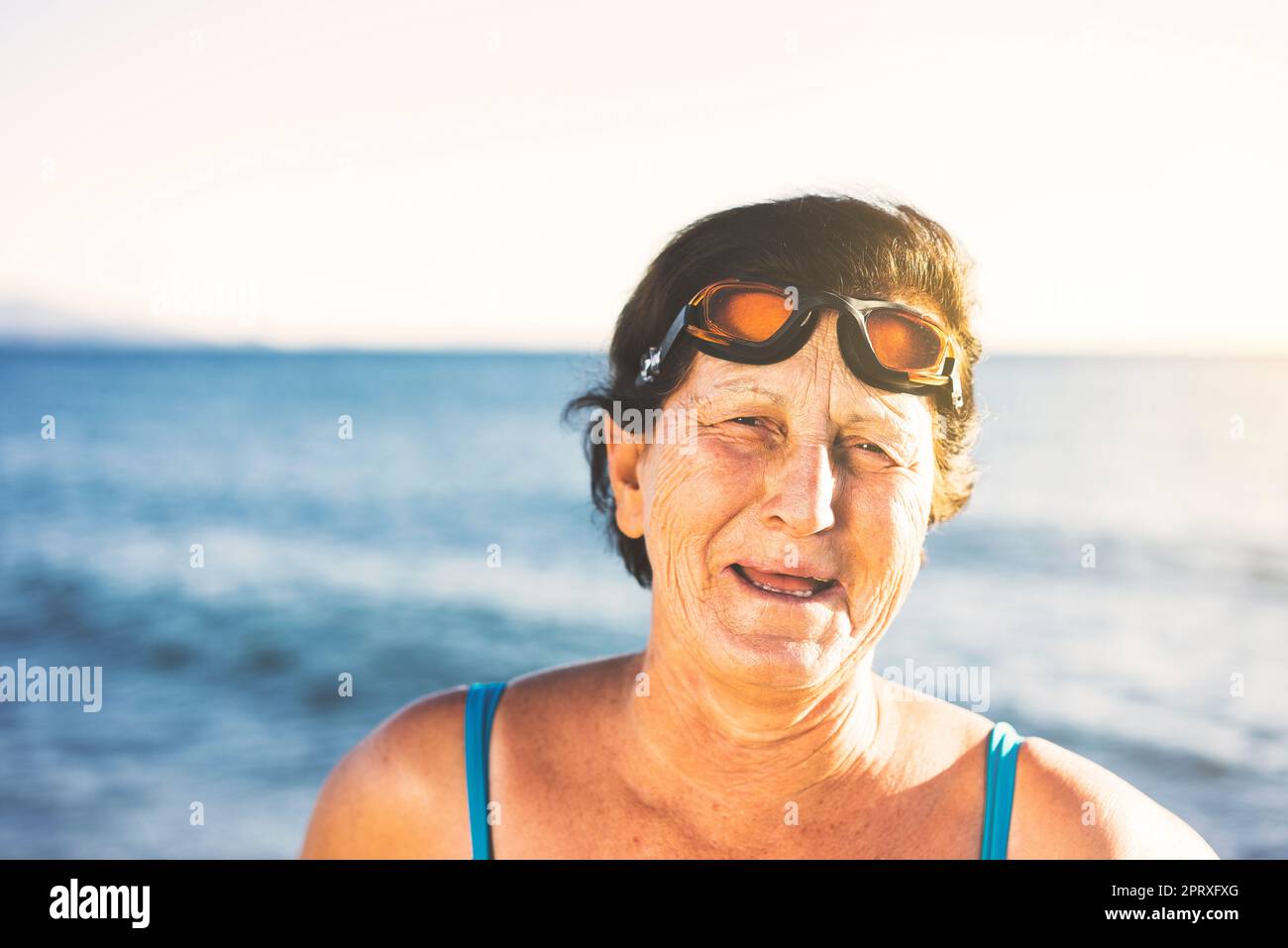 Senior woman at the beach, standing by the sea. Wearing a blue swimsuit ...
